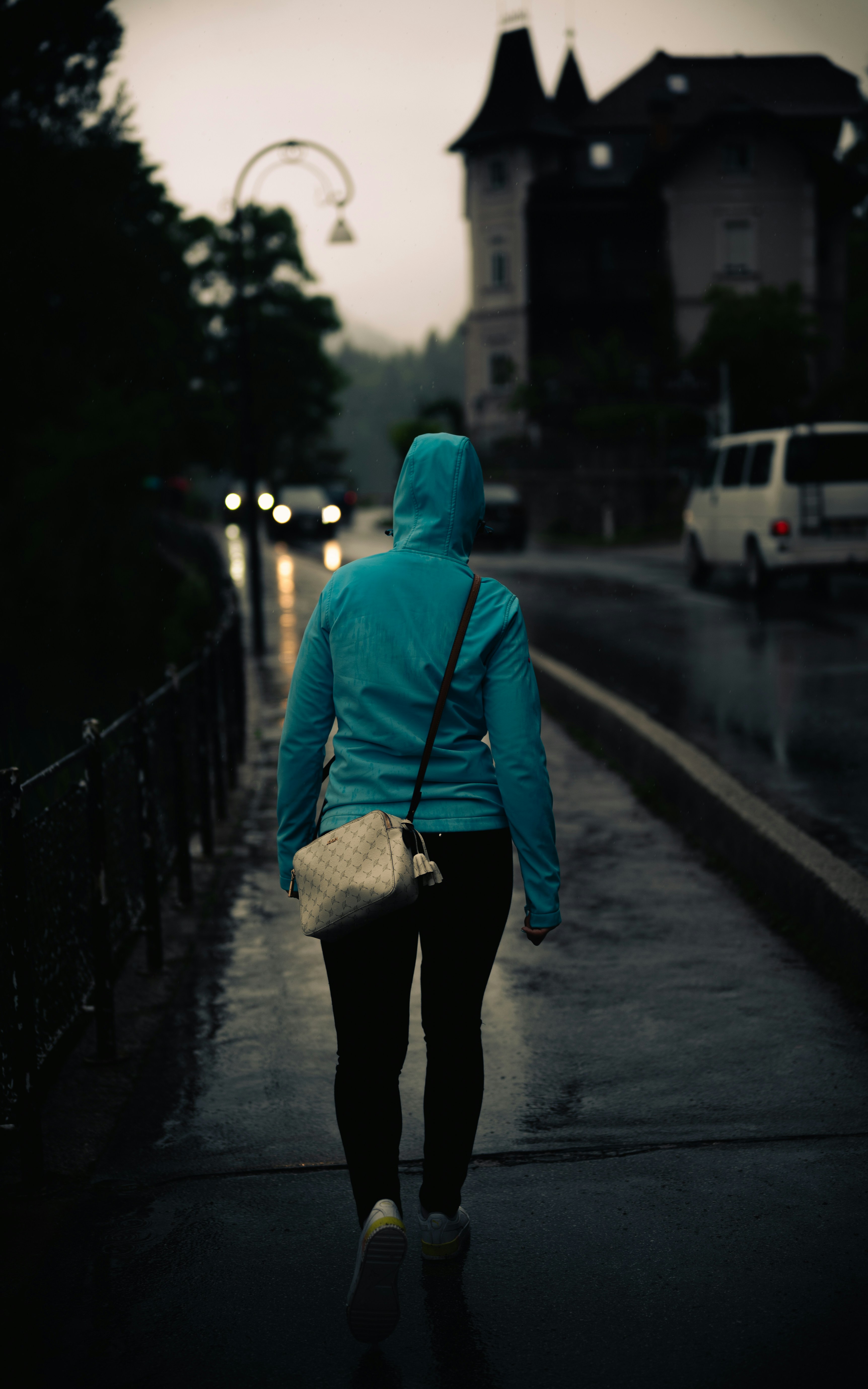 Person in a blue hoodie walking along a rain-soaked street, with blurred cars in the background. The ambiance is moody and reflective.