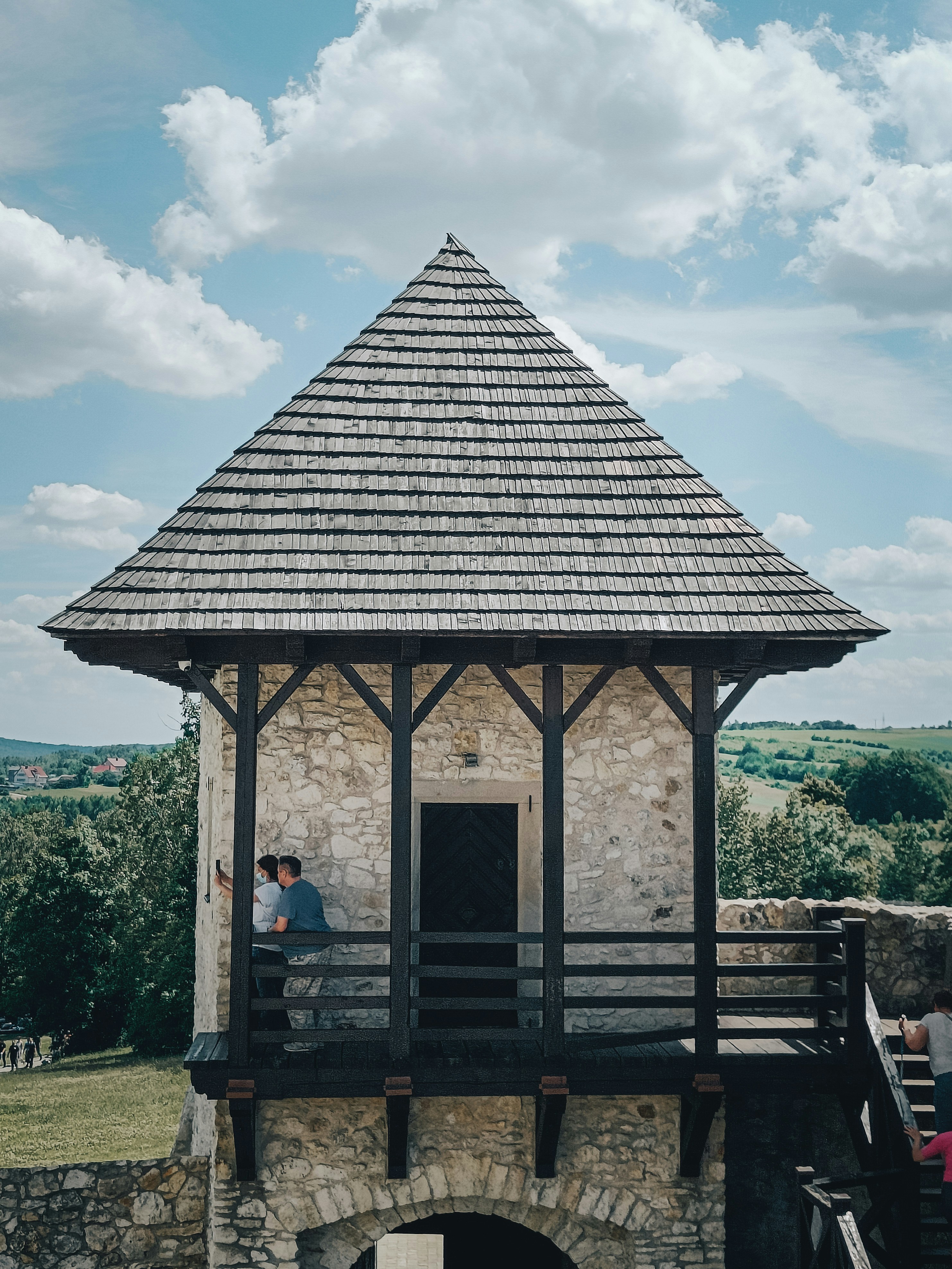 Photograph of a stone watchtower topped with a conical thatched roof and a wooden balcony, set against a rural landscape of fields and trees. Two people stand on the balcony, enjoying the view.