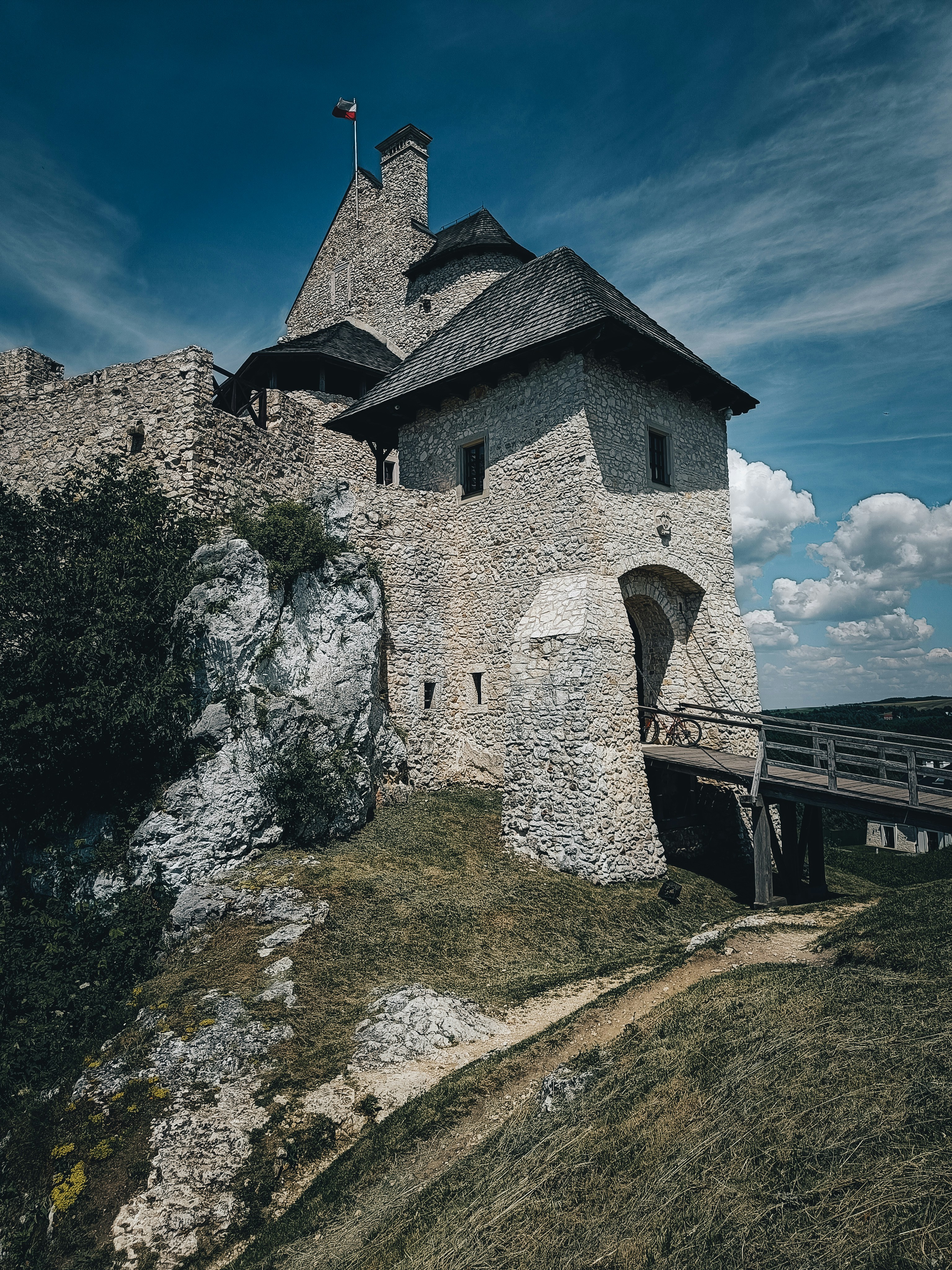 Medieval stone fortress perched on a rocky outcrop, with a wooden bridge leading to its entrance. A flag flutters atop the structure against a backdrop of blue skies.