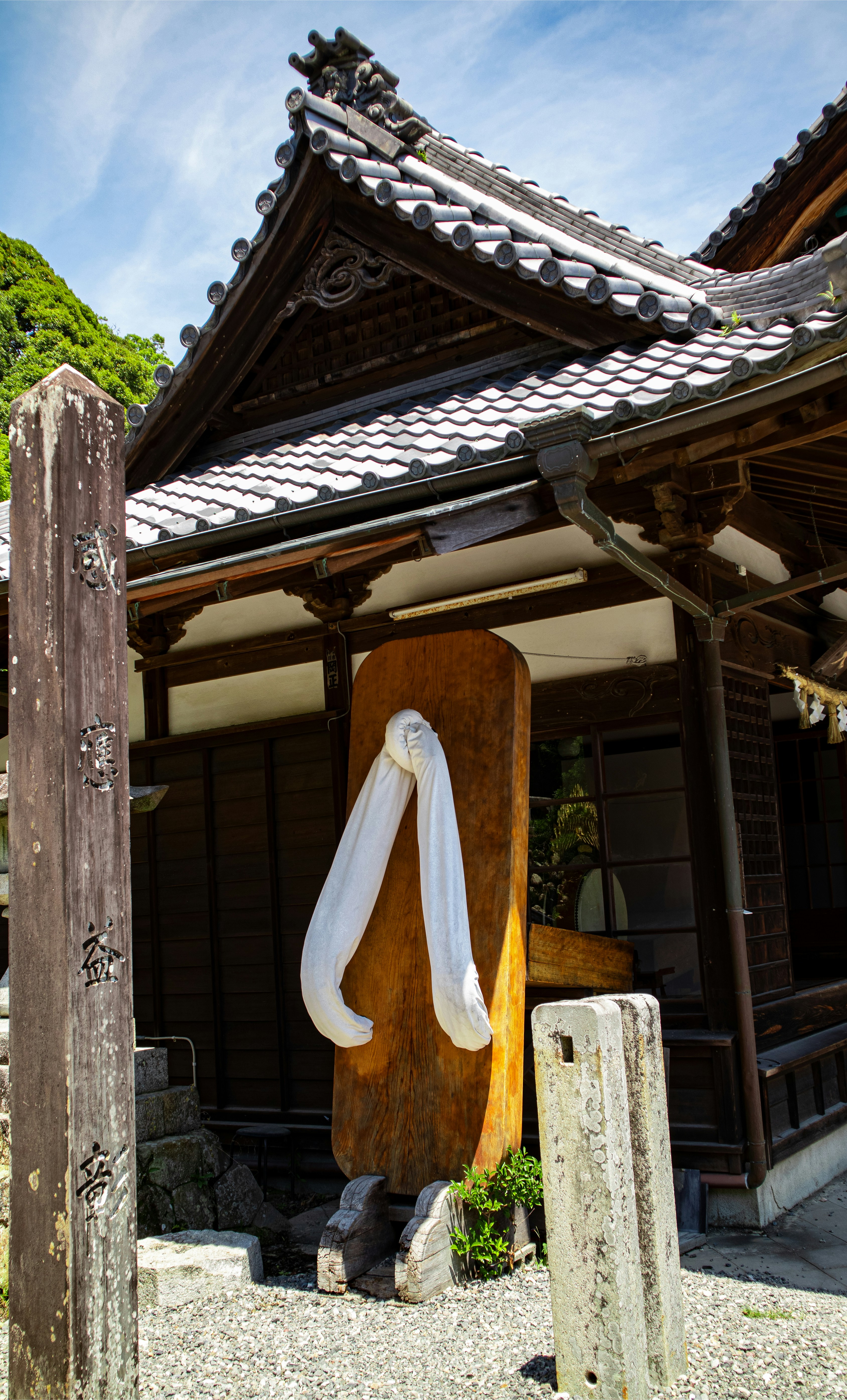A wooden sculpture adorned with a white cloth stands in front of a traditional Japanese building, symbolizing cultural heritage and reverence. The intricate roof and surrounding elements enhance the scene.