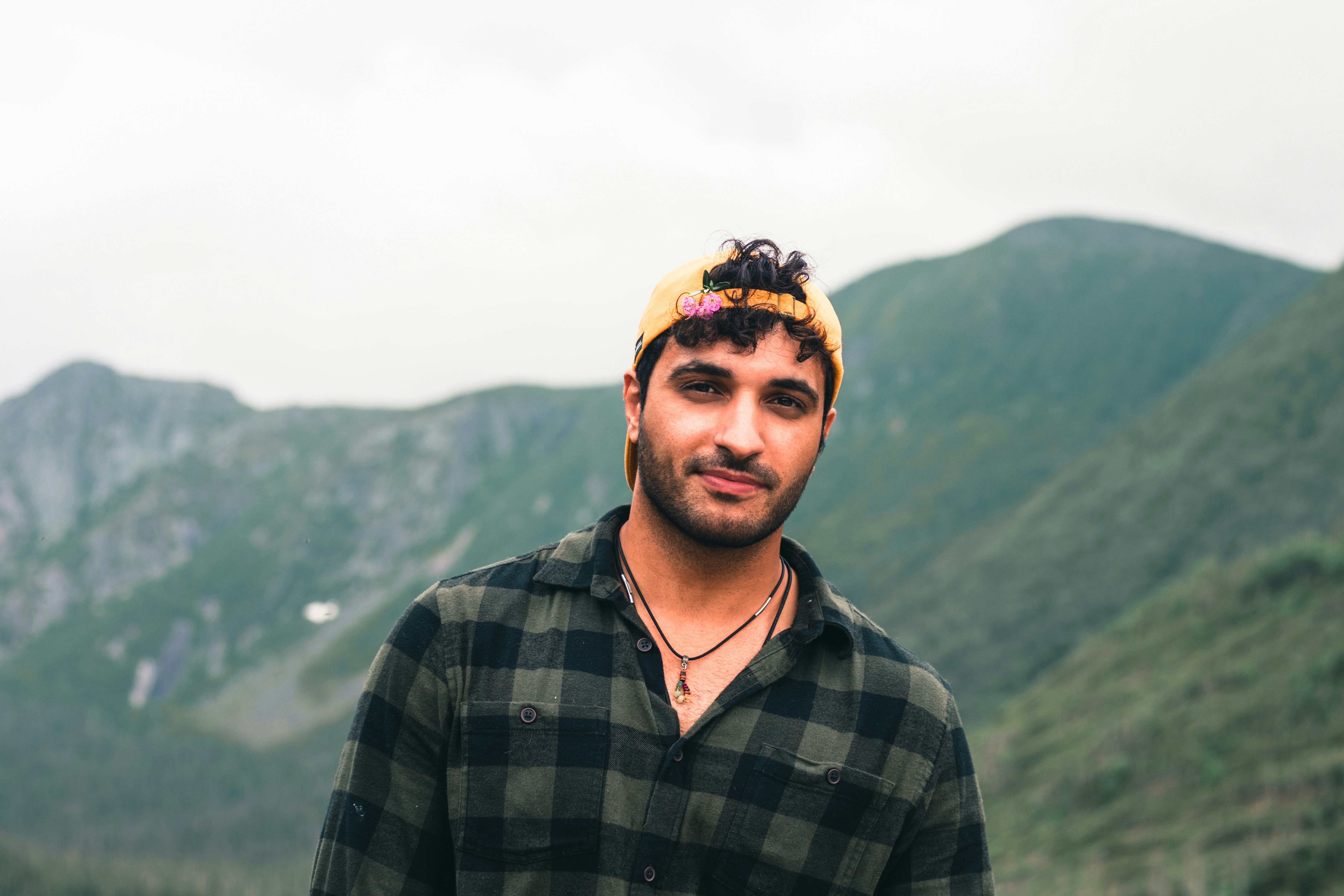 Man in a plaid shirt and hat stands before misty green mountains.