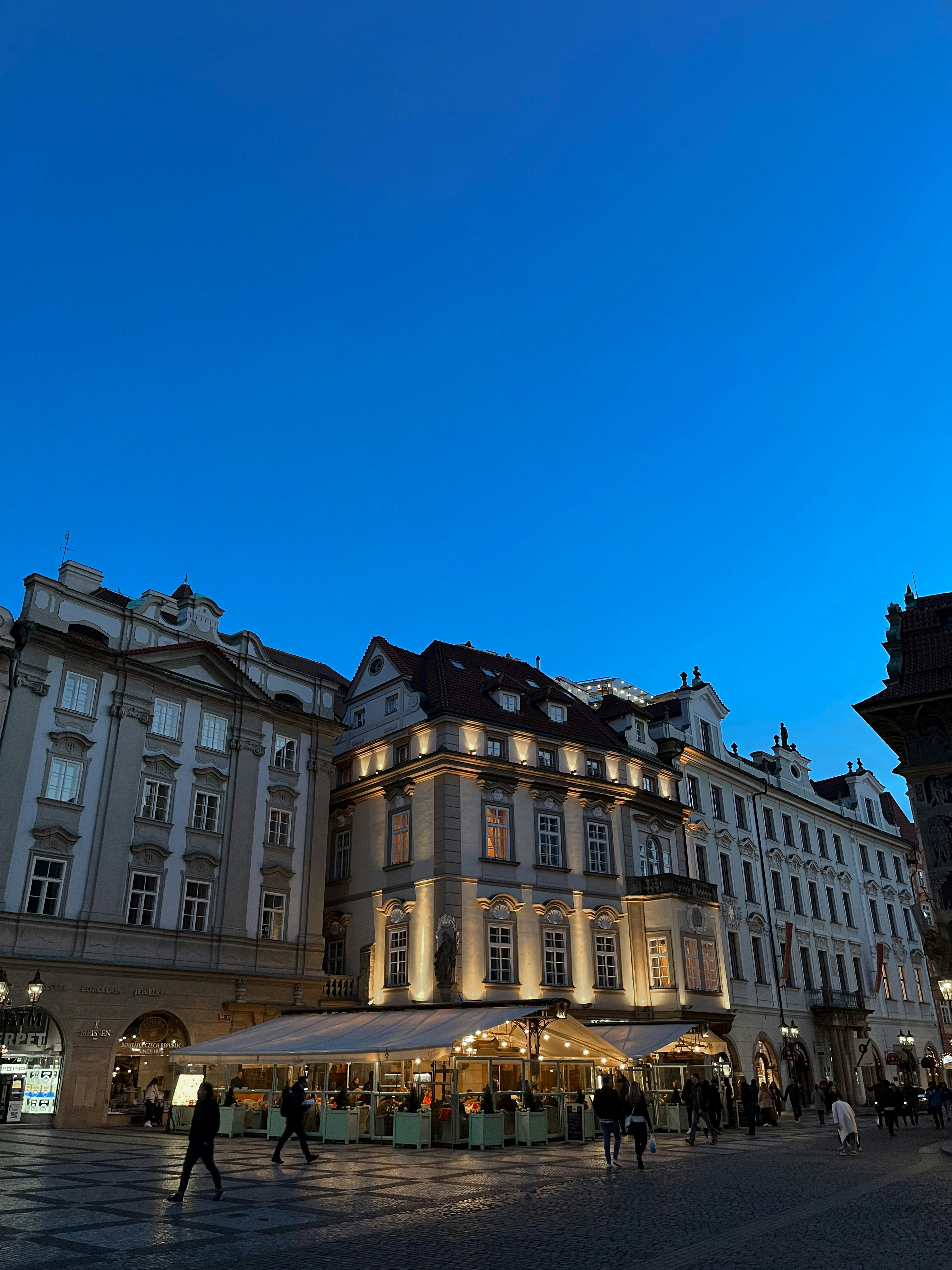 Historic buildings illuminated under a deep blue sky, with people strolling in the foreground. A charming outdoor café adds a lively touch.