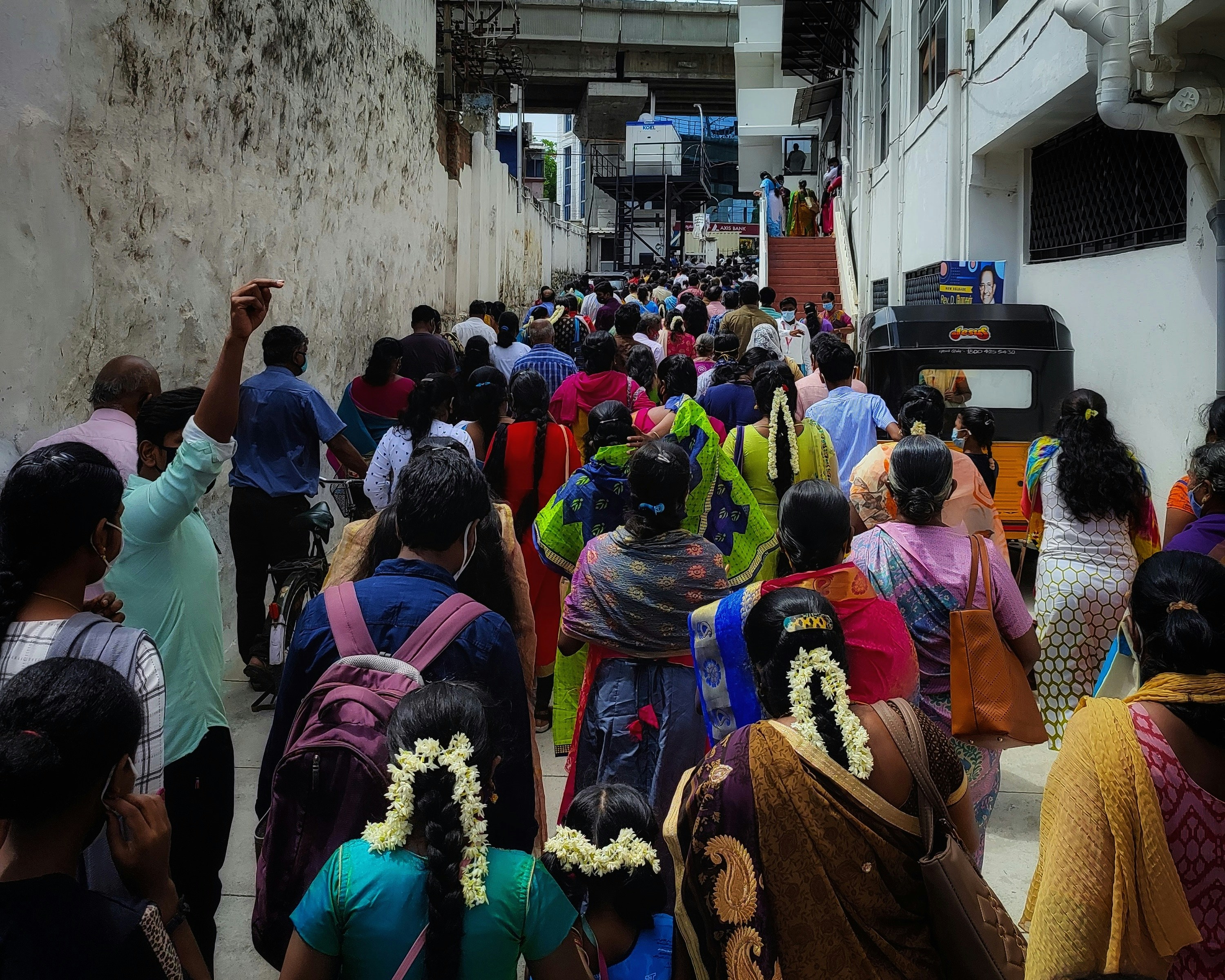 Crowd of people in colorful attire moving through a narrow alley, showcasing a festive atmosphere. Floral decorations adorn the hair of many participants.