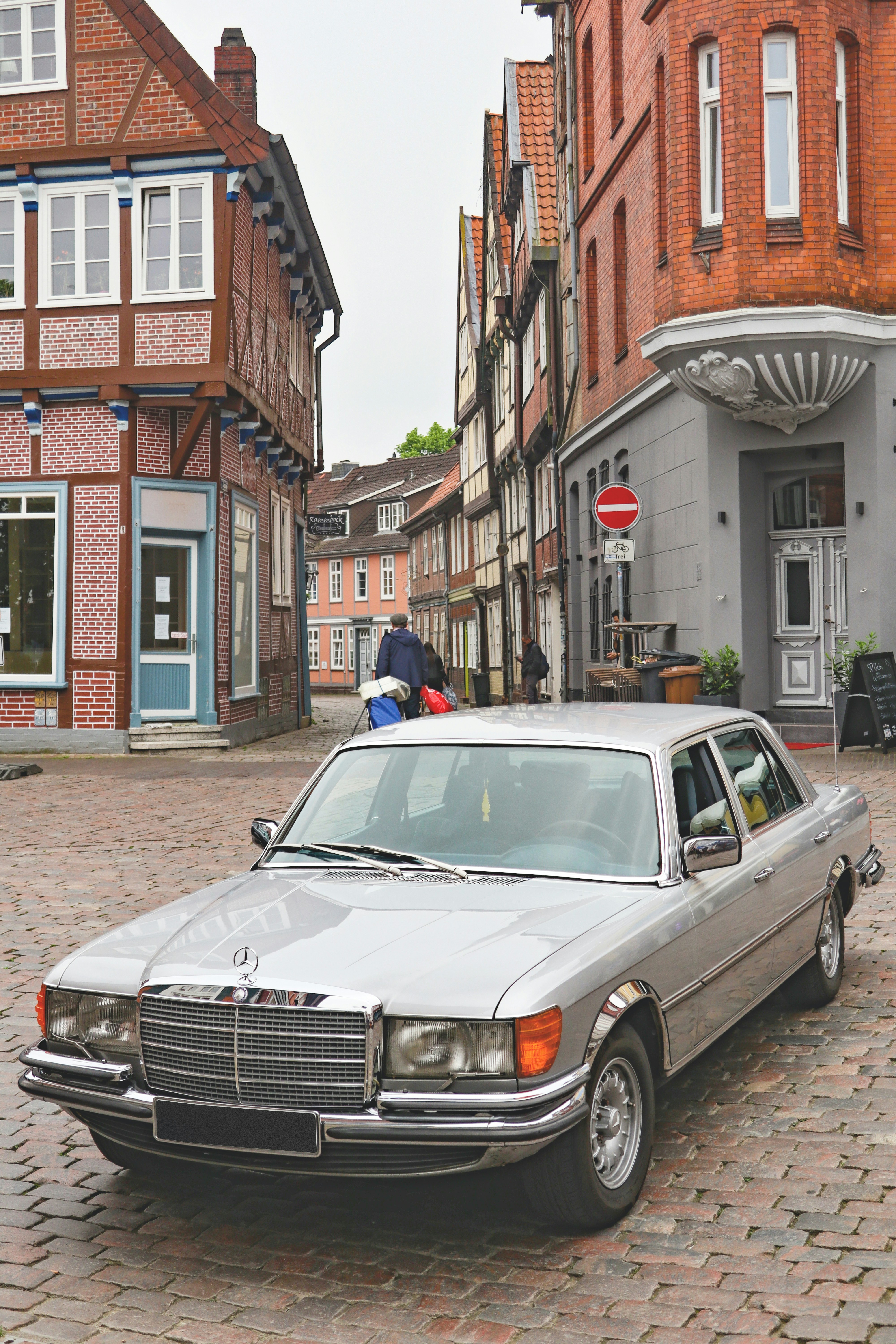Photograph of a vintage Mercedes parked on a cobbled street, framed by brick and timber-framed facades, with pedestrians in the distance.