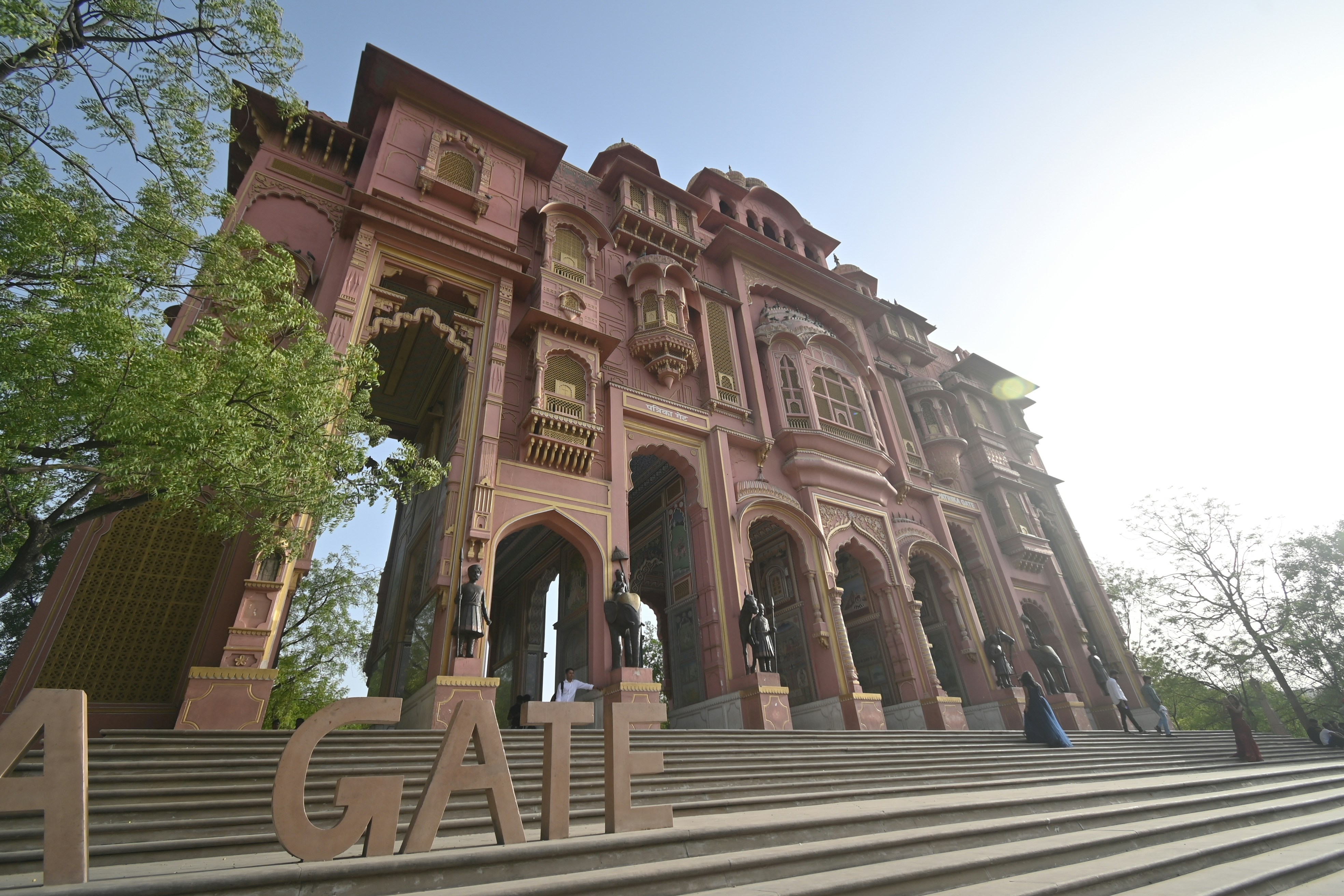 Intricate pink facade of a grand gate with sculptures, framed by lush greenery and a clear sky.