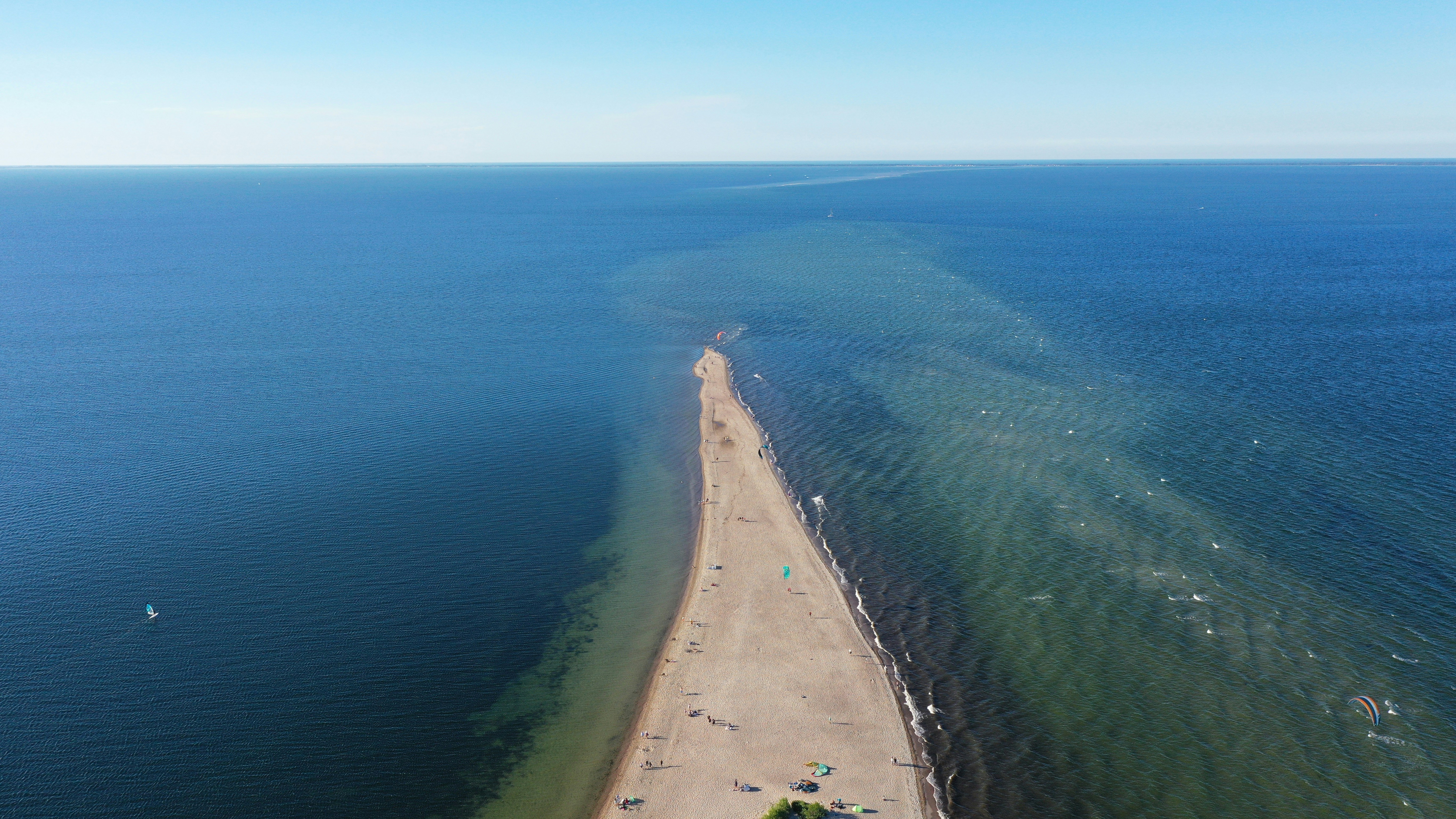 Aerial view of a narrow sandy spit extending into the blue sea under a clear sky.