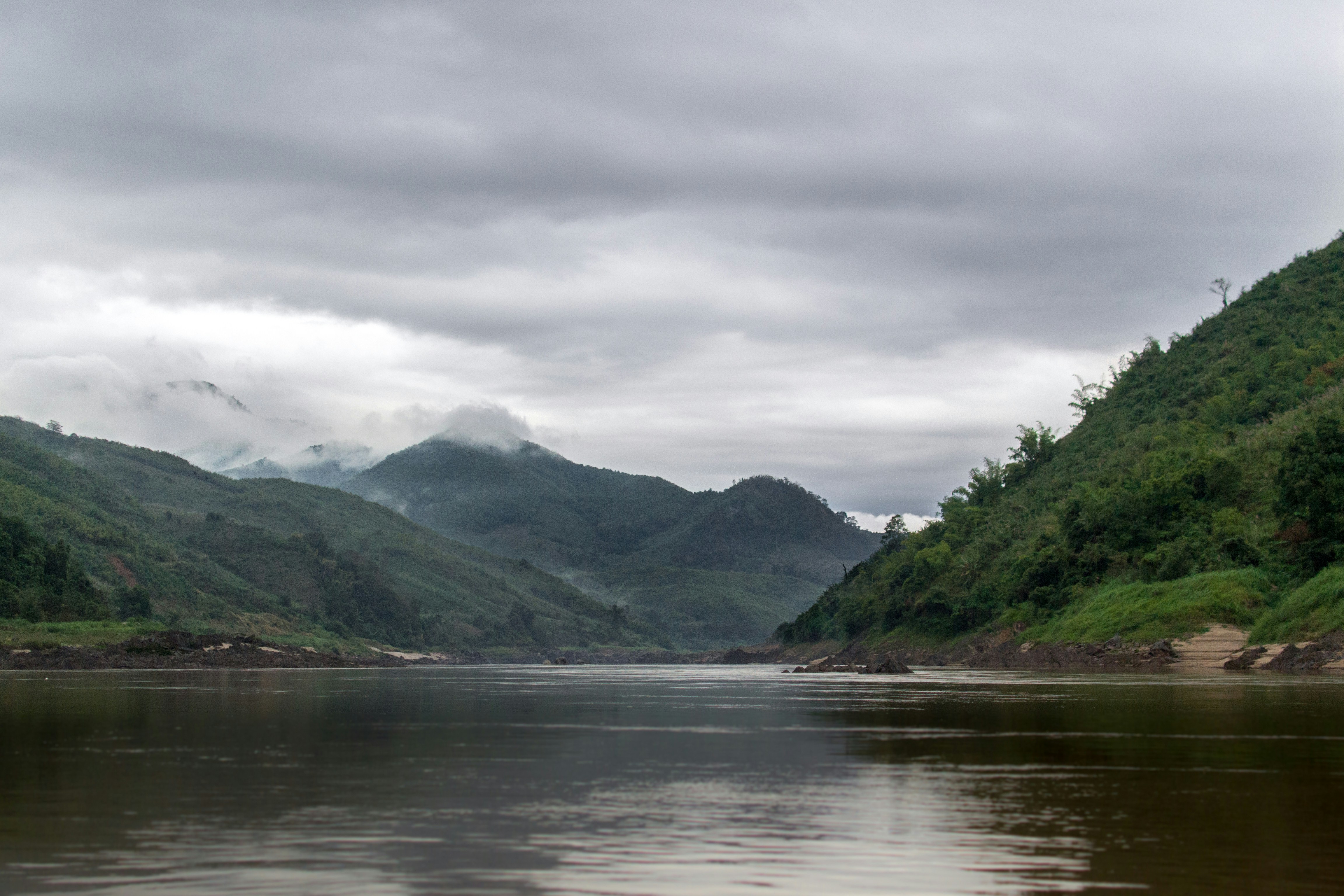 Cloud-shrouded mountains rise above a calm river reflecting the overcast sky.