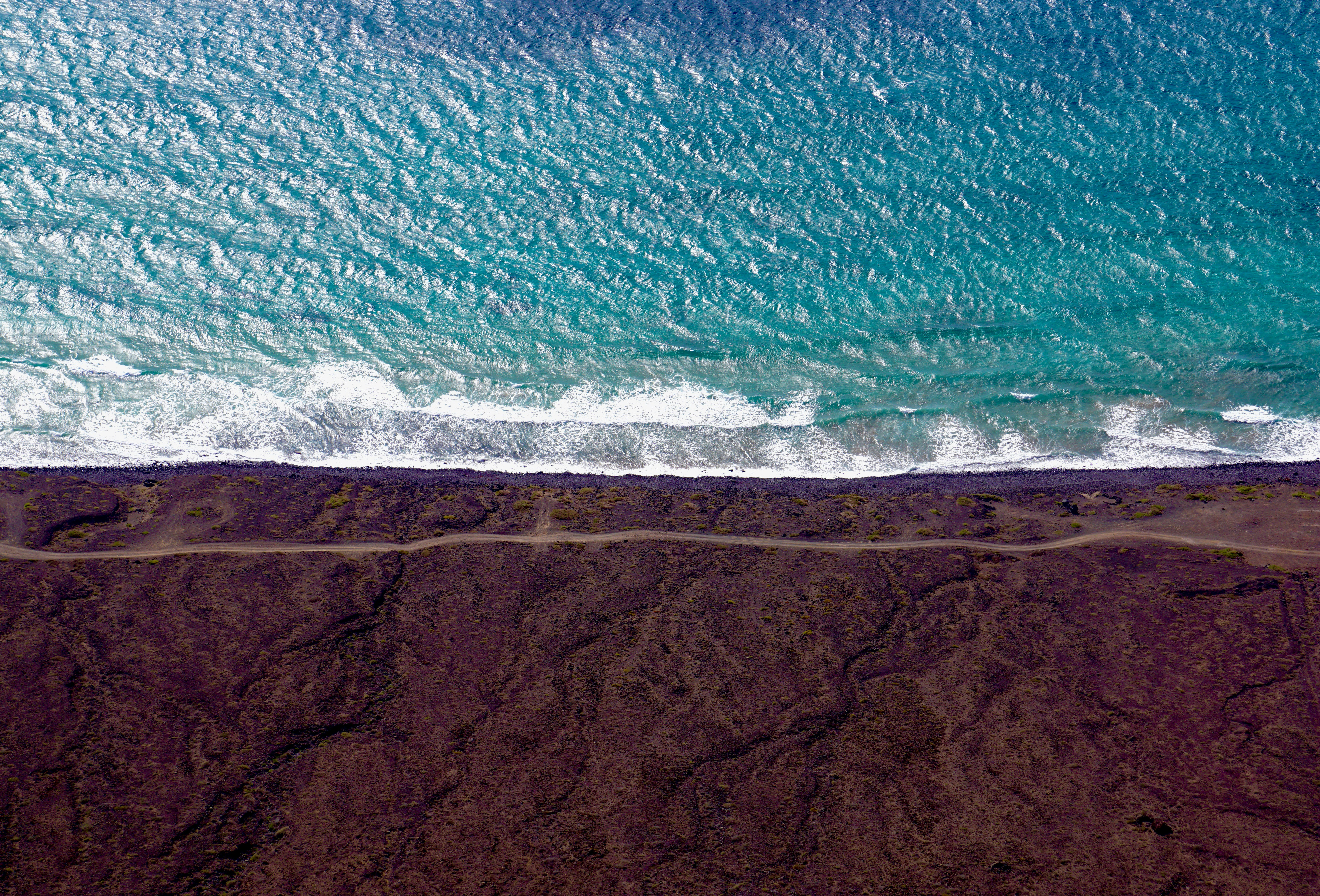 Aerial view showcasing the vibrant turquoise ocean waves crashing against a rugged shoreline, with a contrasting dark land texture. 