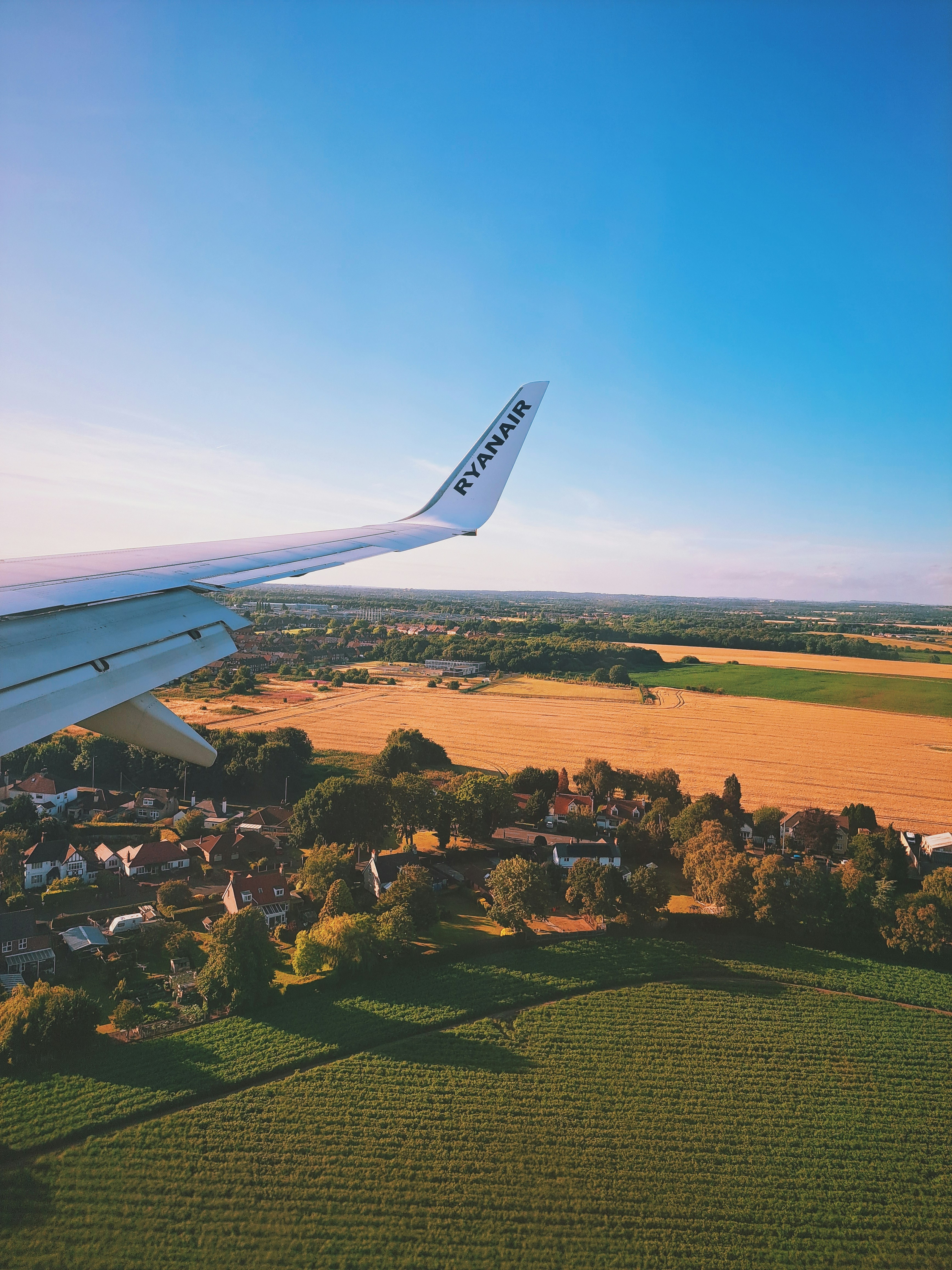 Airplane wing extending over vibrant farmland and quaint houses during descent. Clear blue sky enhances the scene.