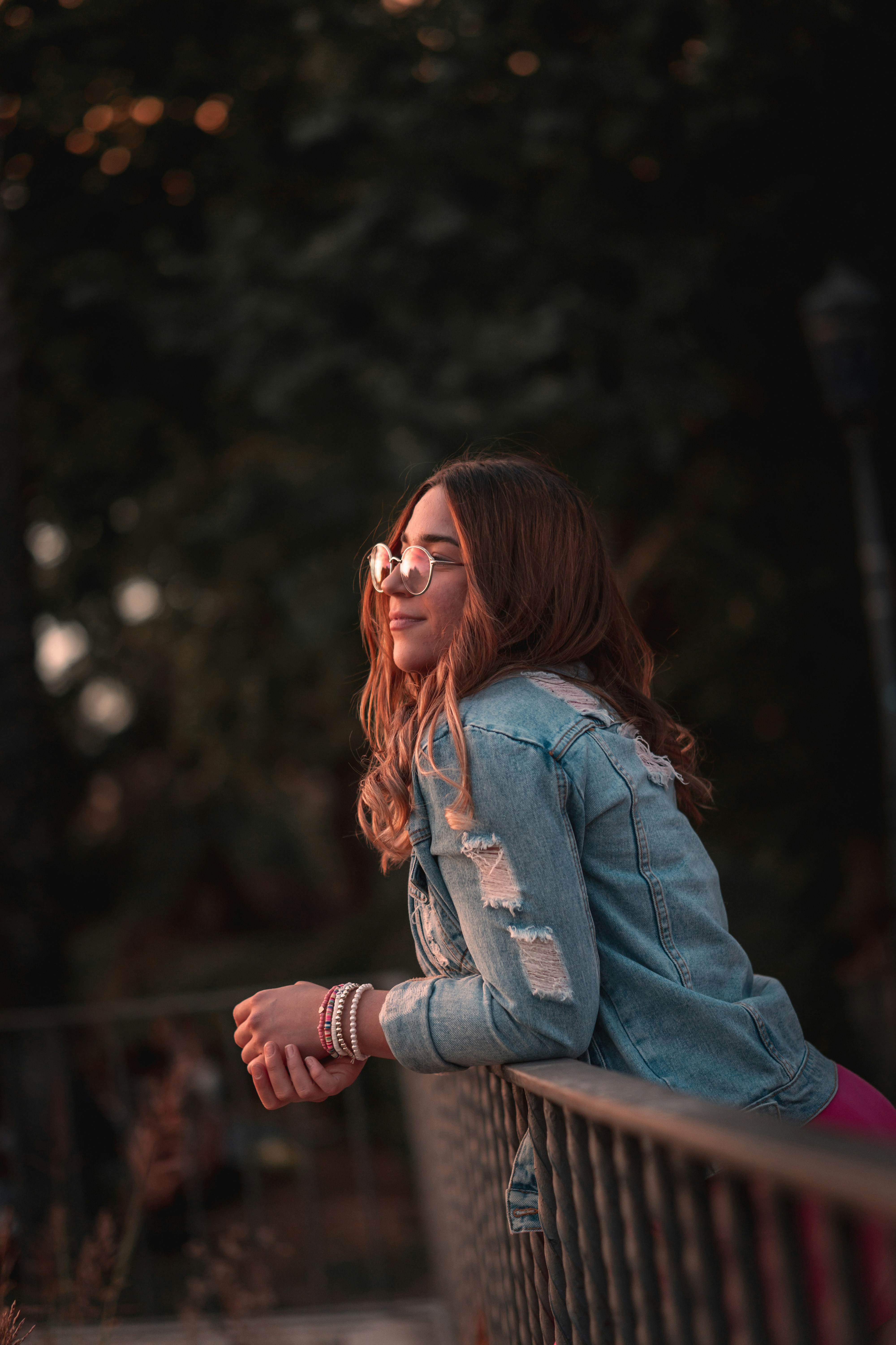 Young woman in a denim jacket leaning on a railing, gazing thoughtfully into the distance during sunset.