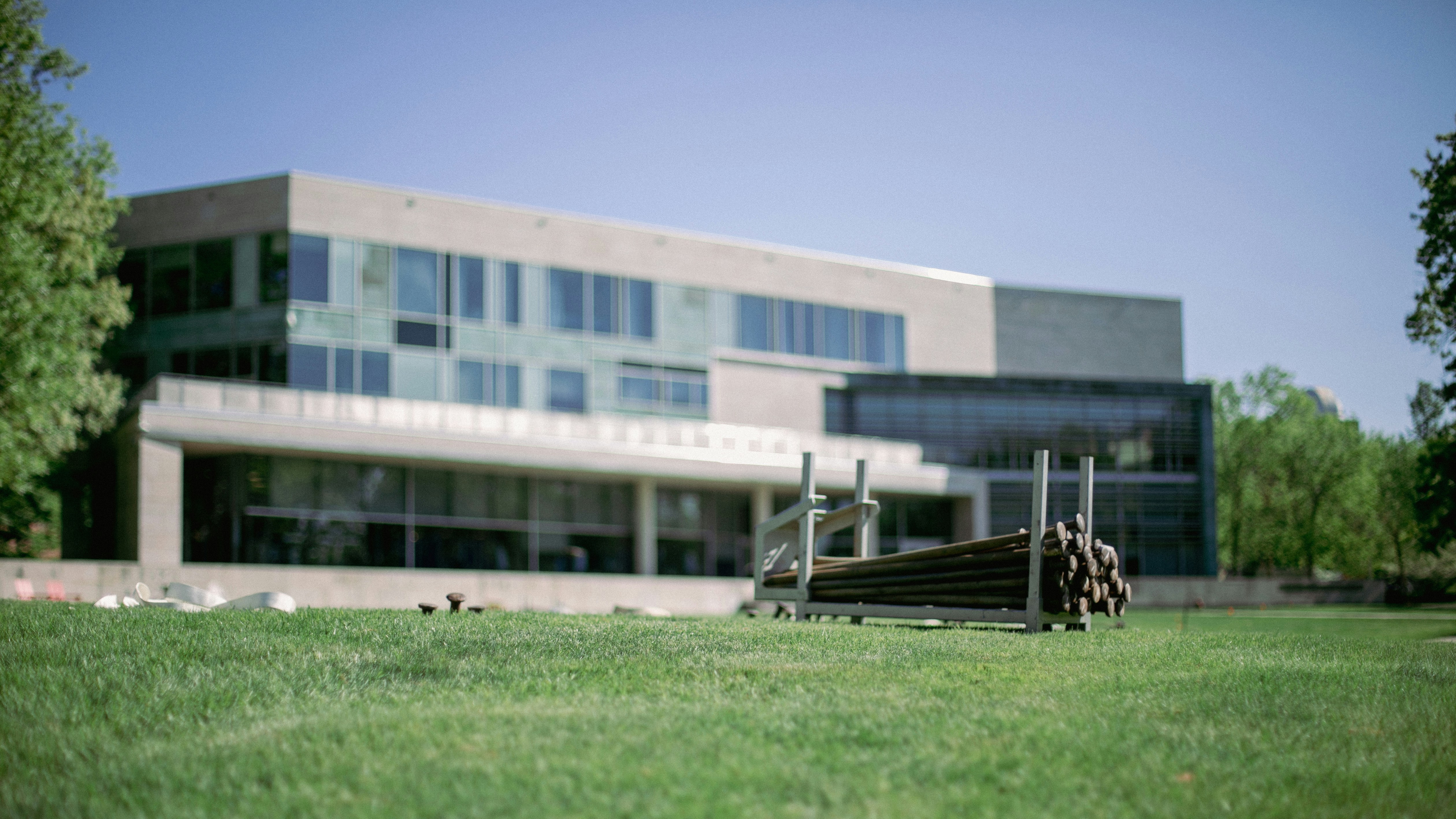 Stack of metal pipes on green grass with a contemporary building in the background, showcasing a blend of urban design and natural elements.