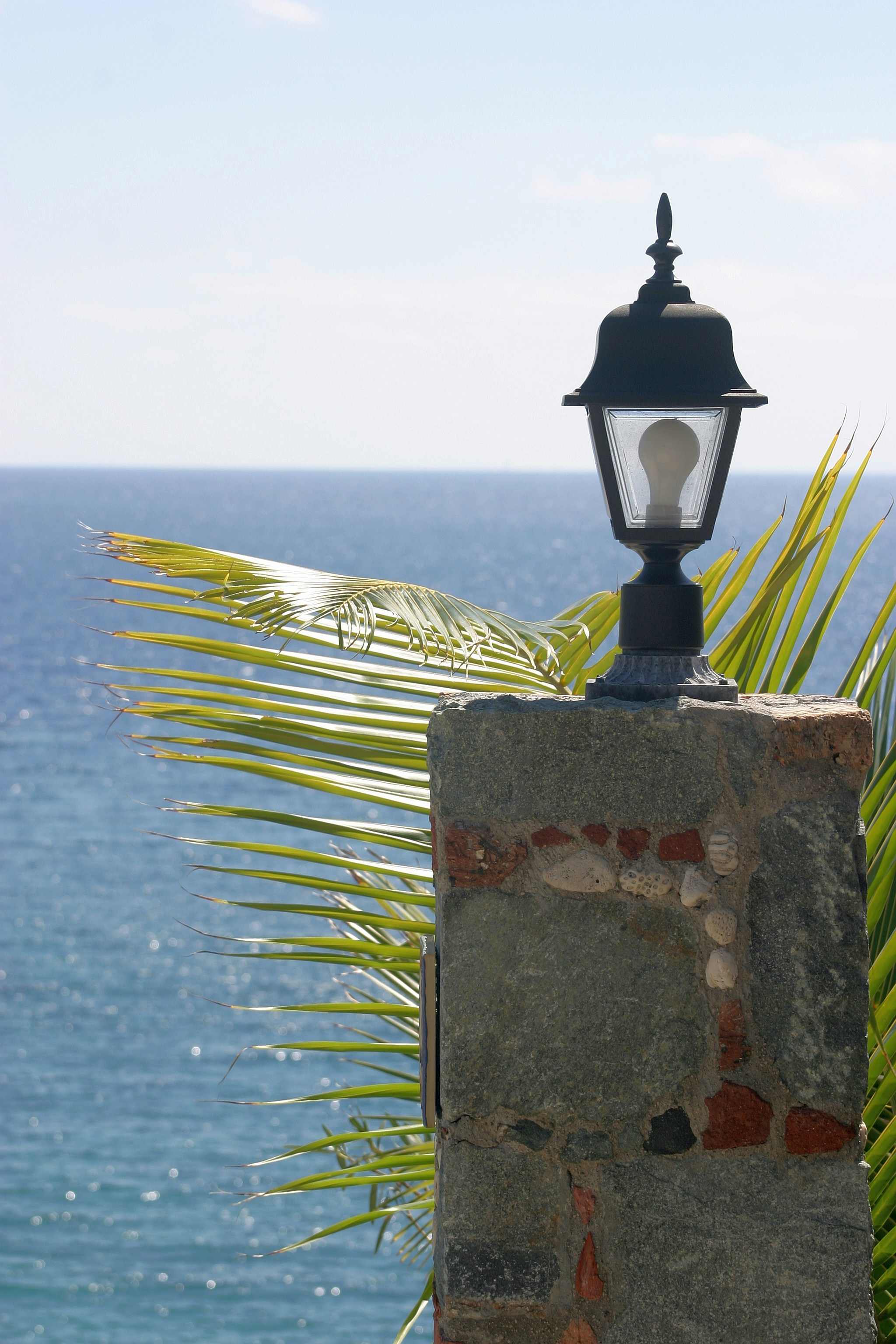 Vintage lamp post atop a stone wall with palm fronds swaying in the breeze, overlooking a tranquil sea.