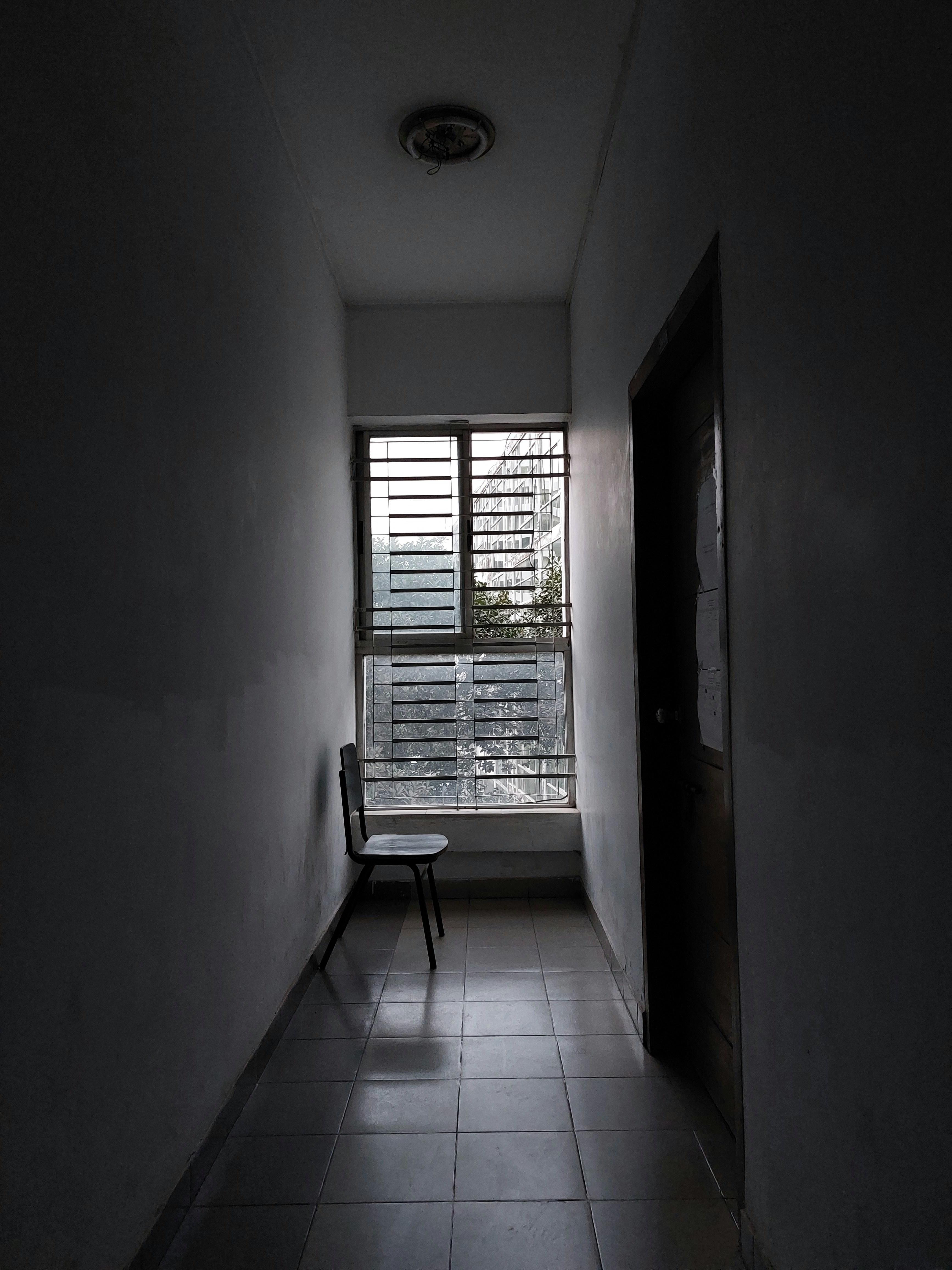 Empty chair positioned in a narrow hallway, illuminated by natural light from a window. The stark contrast highlights the solitude of the scene.