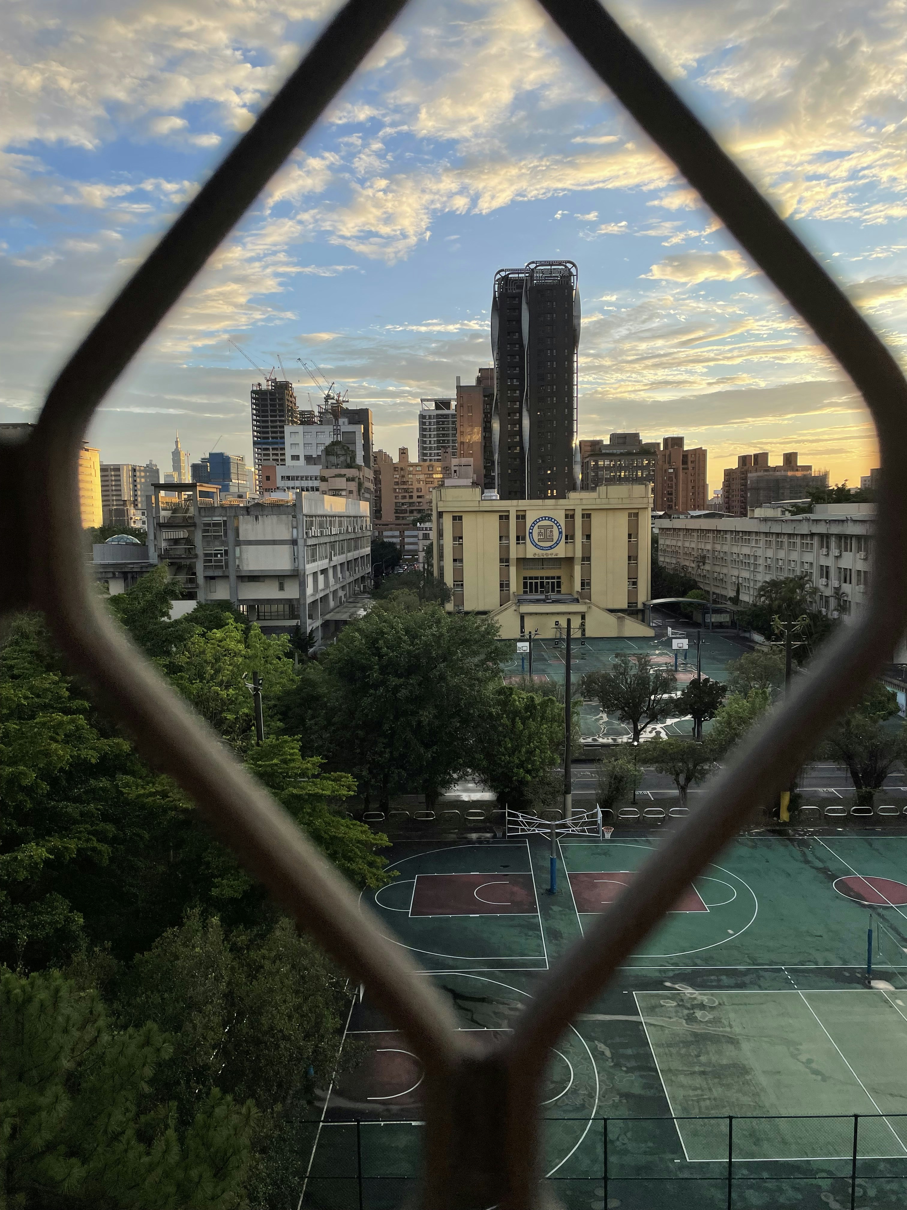 View of a city skyline framed by a diamond-patterned fence, showcasing a blend of modern architecture and greenery. The scene captures the essence of urban life at dusk.
