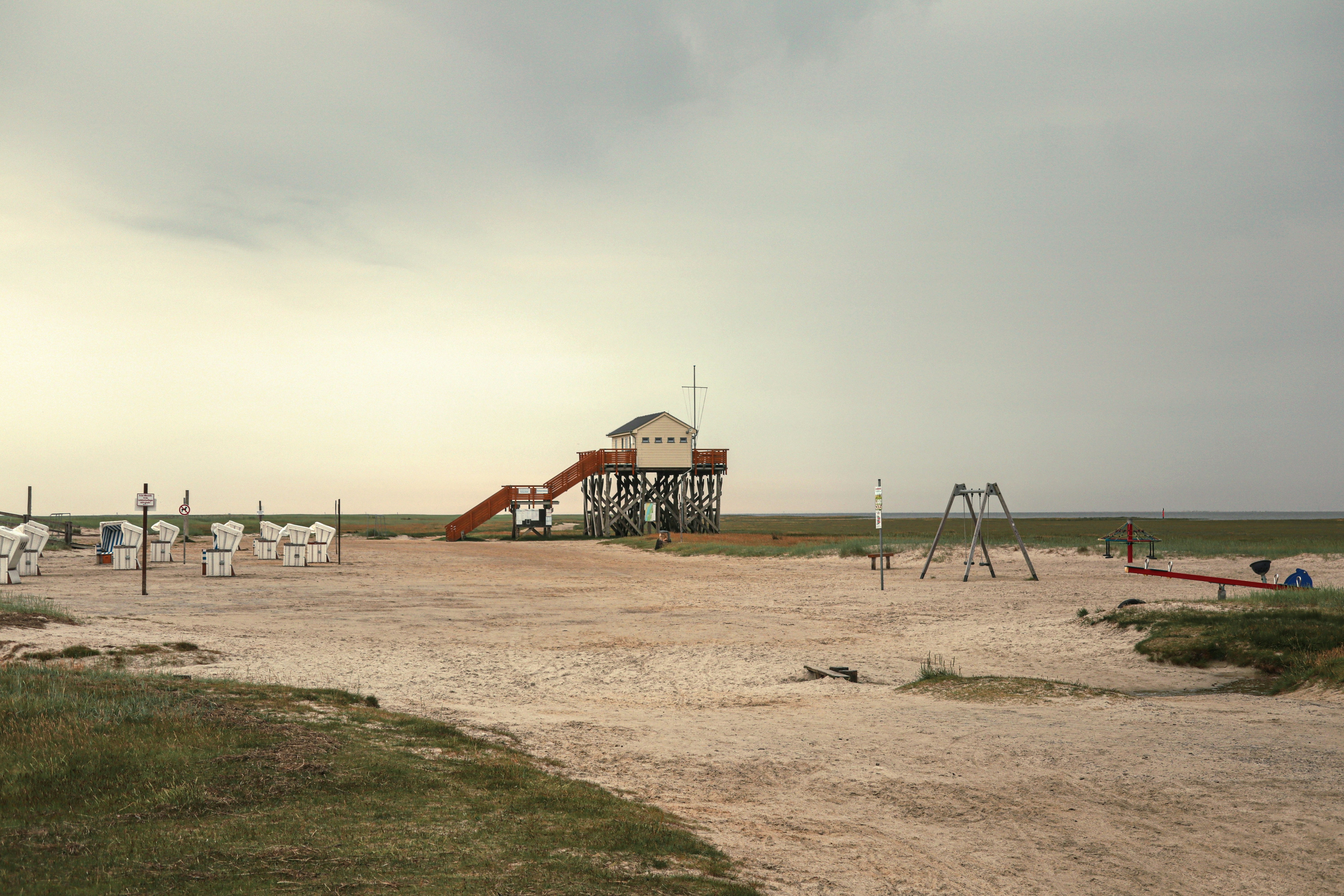 A wooden observation tower stands on stilts above a sandy beach, surrounded by empty chairs and playground equipment. The scene captures a tranquil coastal atmosphere under an overcast sky.