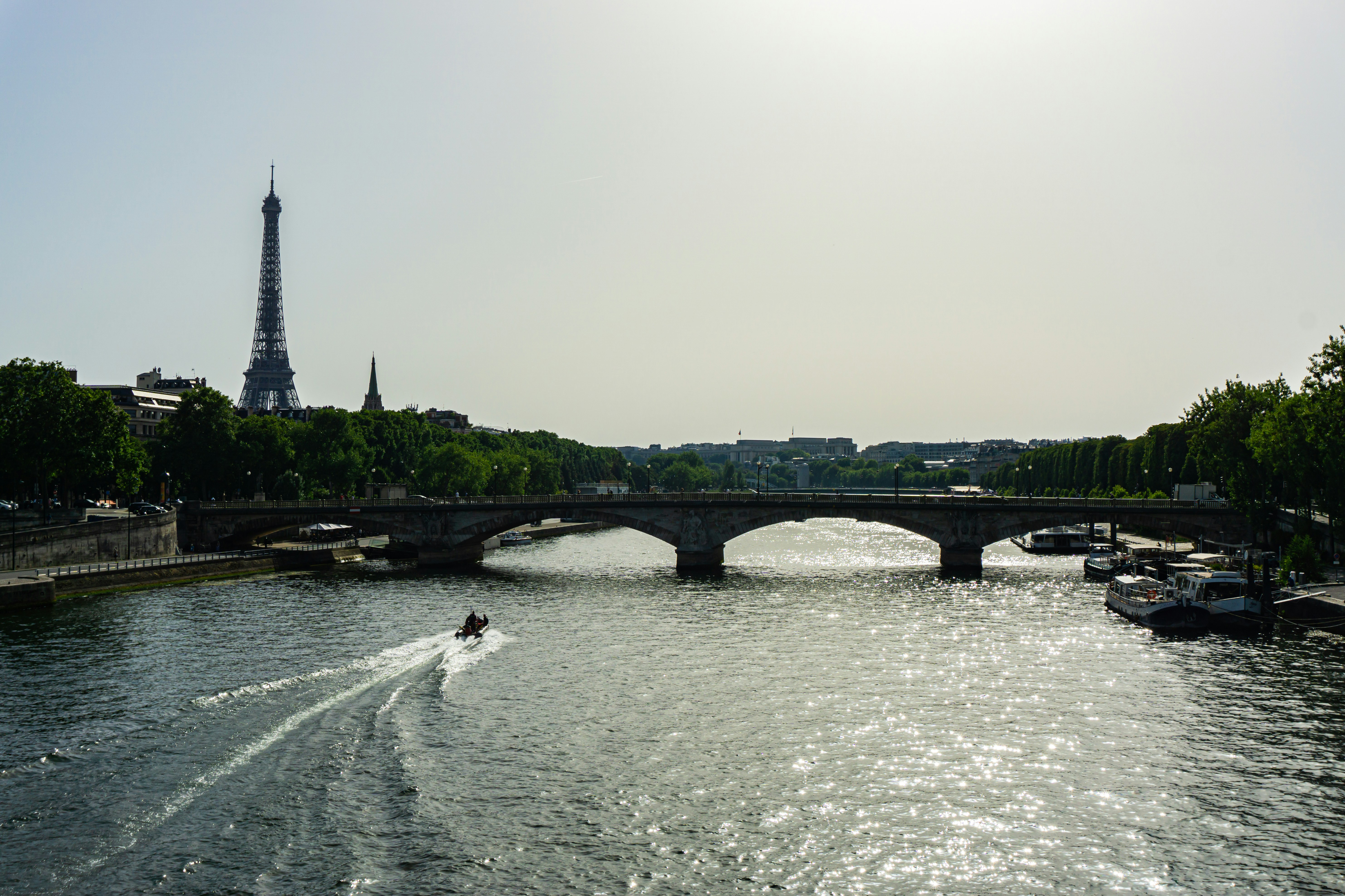 A speedboat glides across the Seine River, framed by the iconic silhouette of the Eiffel Tower and lush riverside greenery.