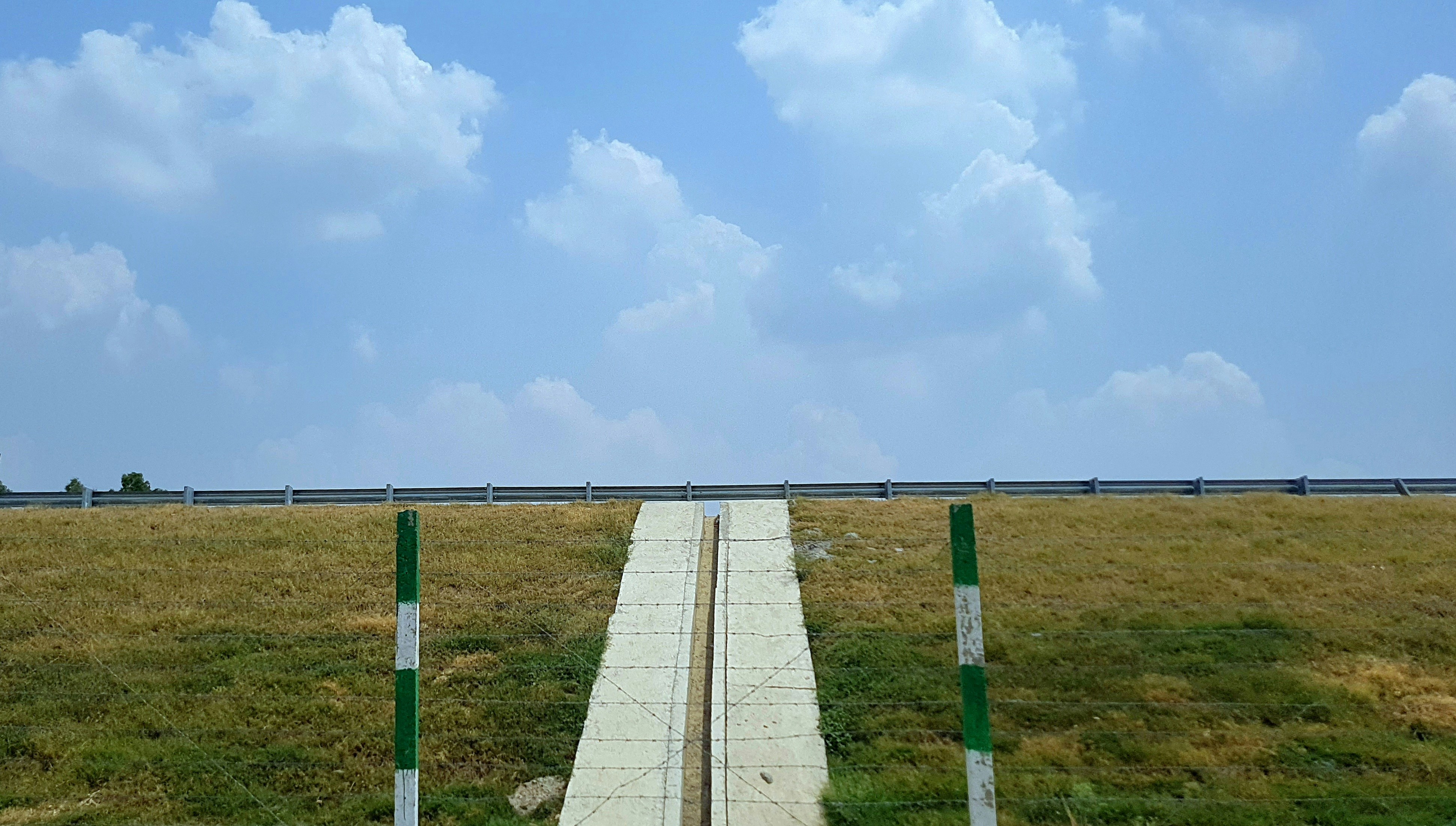 Concrete pathway leading towards open fields under a blue sky dotted with clouds.
