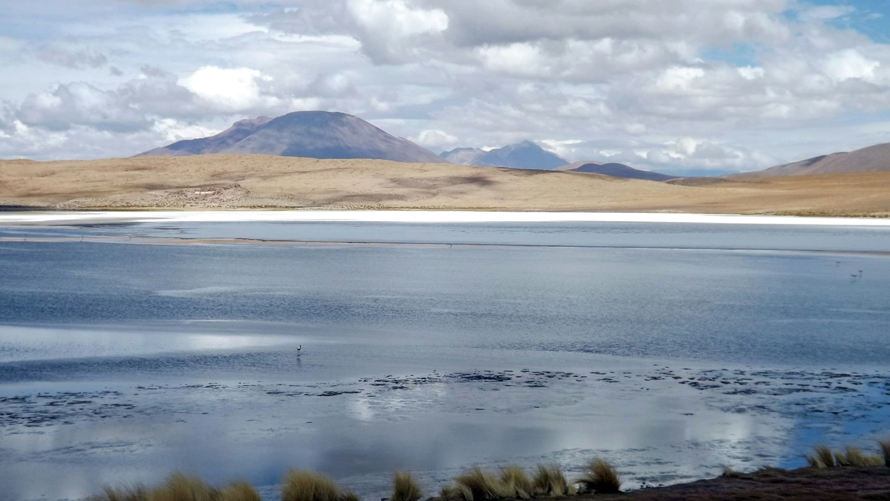 Expansive view of a tranquil high-altitude lake reflecting the sky, surrounded by distant mountains and arid land.