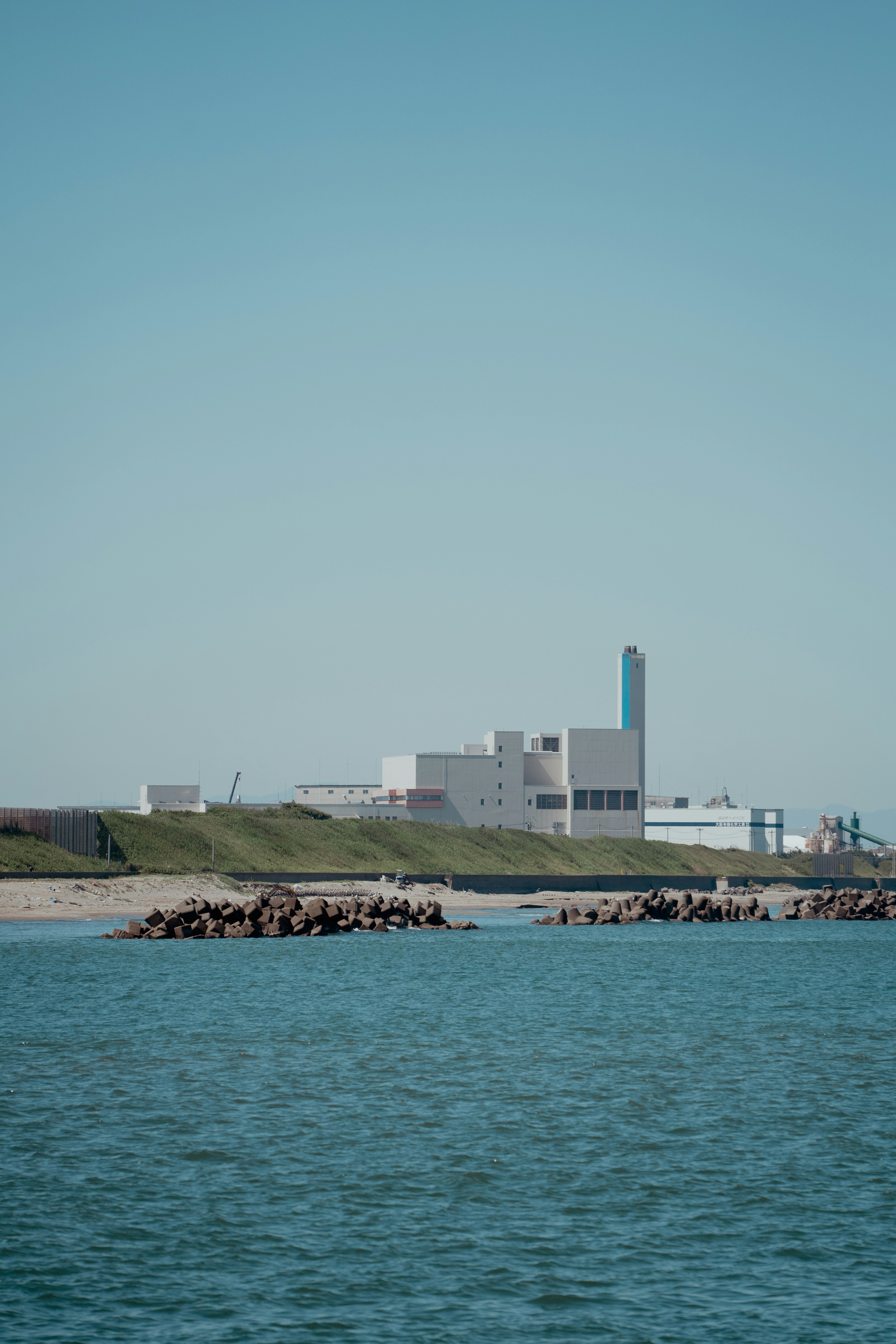 A coastal industrial facility stands against a clear blue sky, framed by gentle waves and rocky formations in the foreground.