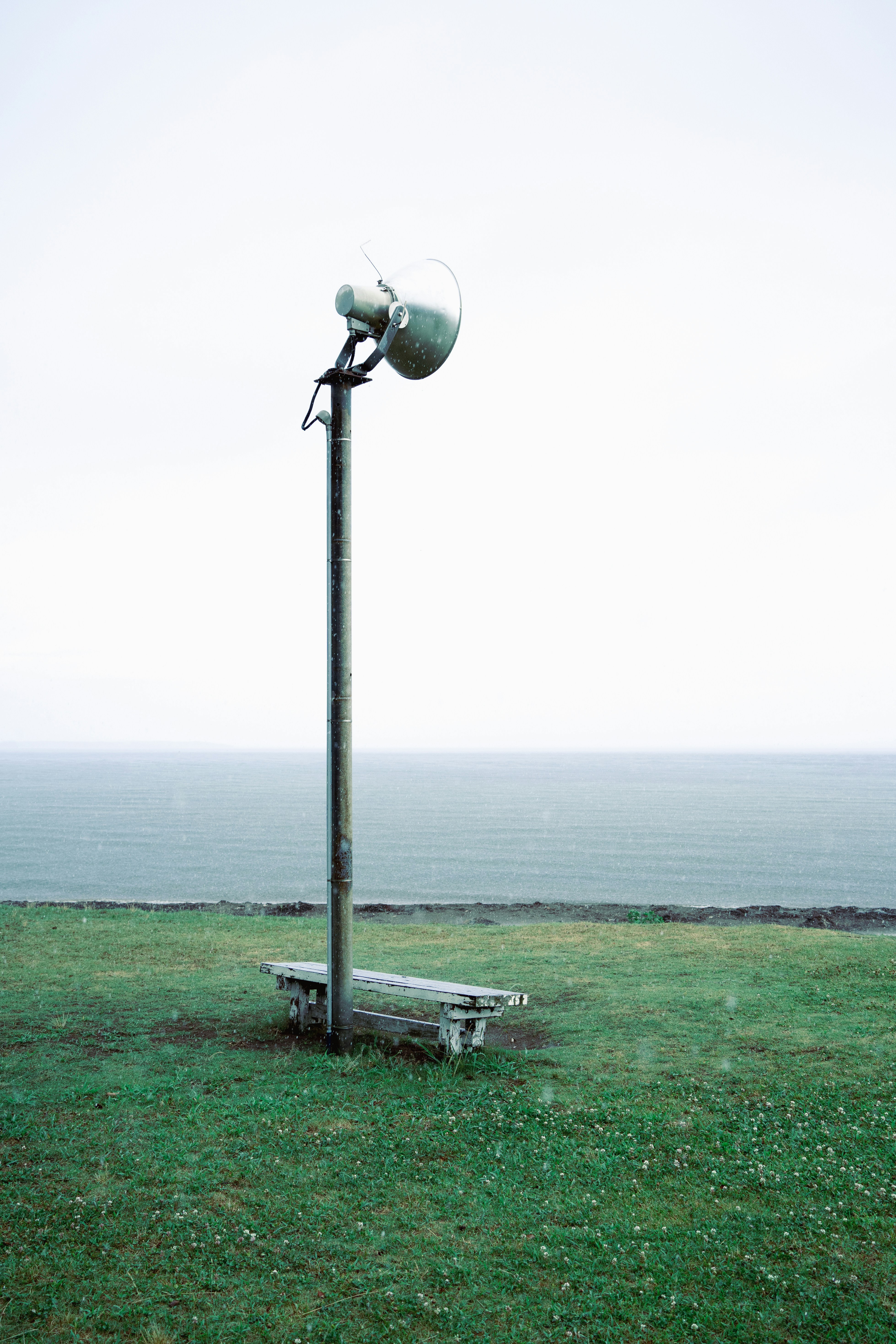 A weathered metal pole with a round dish rises beside a simple wooden bench on grassy coastal ground, looking toward the calm sea. Pale sky forms a near-blank backdrop.