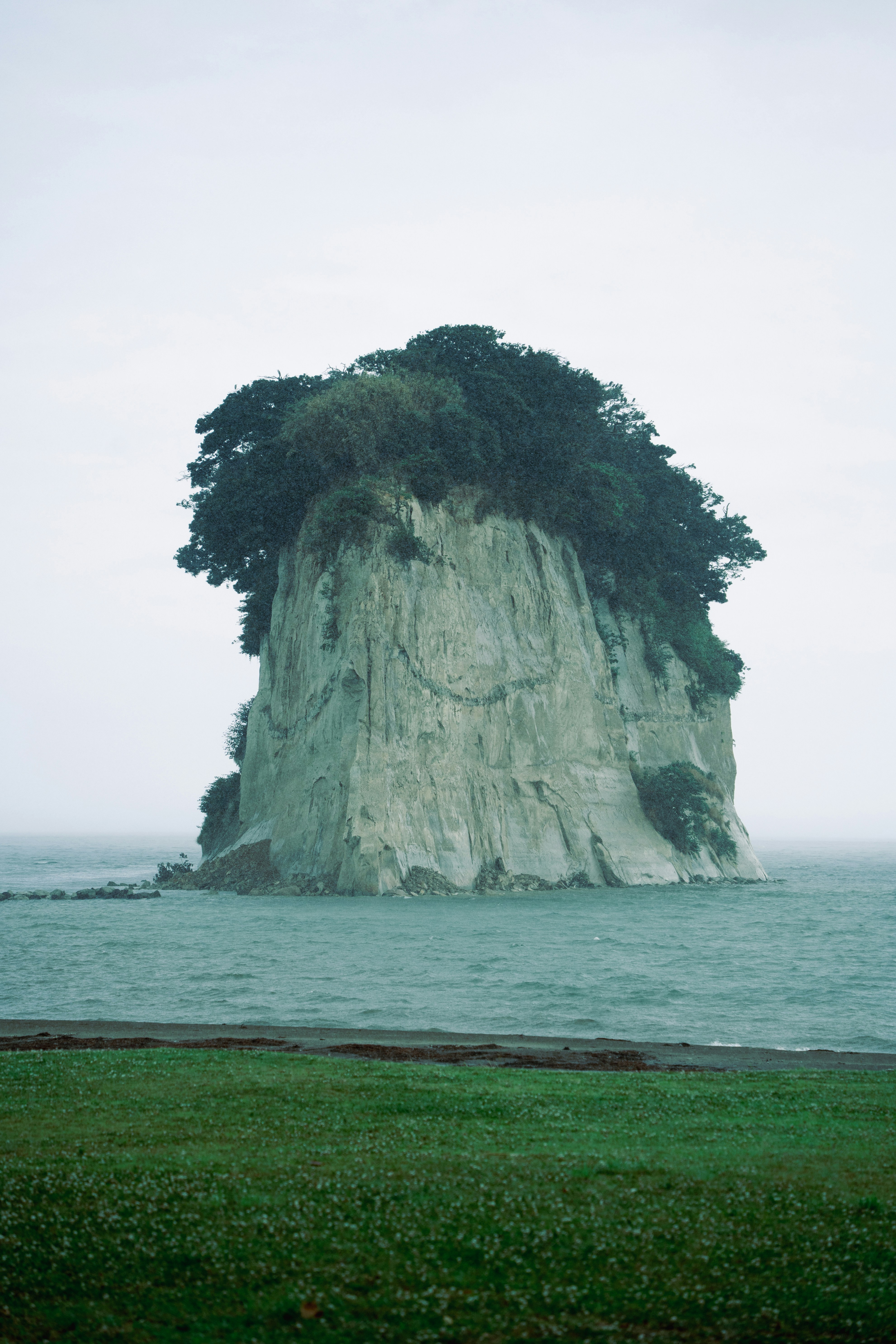 A towering rock formation adorned with lush greenery rises dramatically from the ocean, shrouded in misty weather.