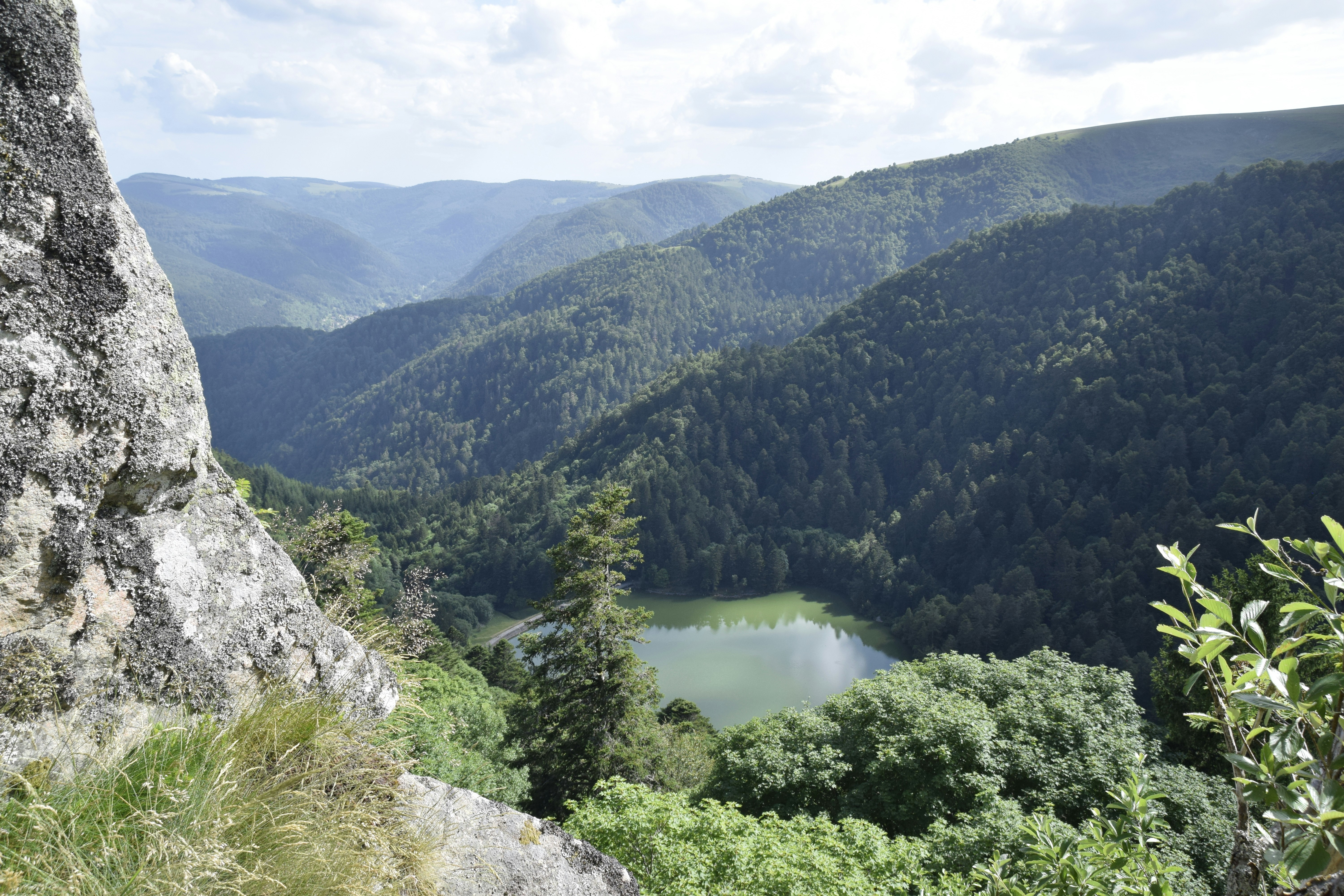 Lush green mountains frame a serene lake nestled in the valley, with rocky ledges in the foreground. A clear sky adds to the tranquil atmosphere.
