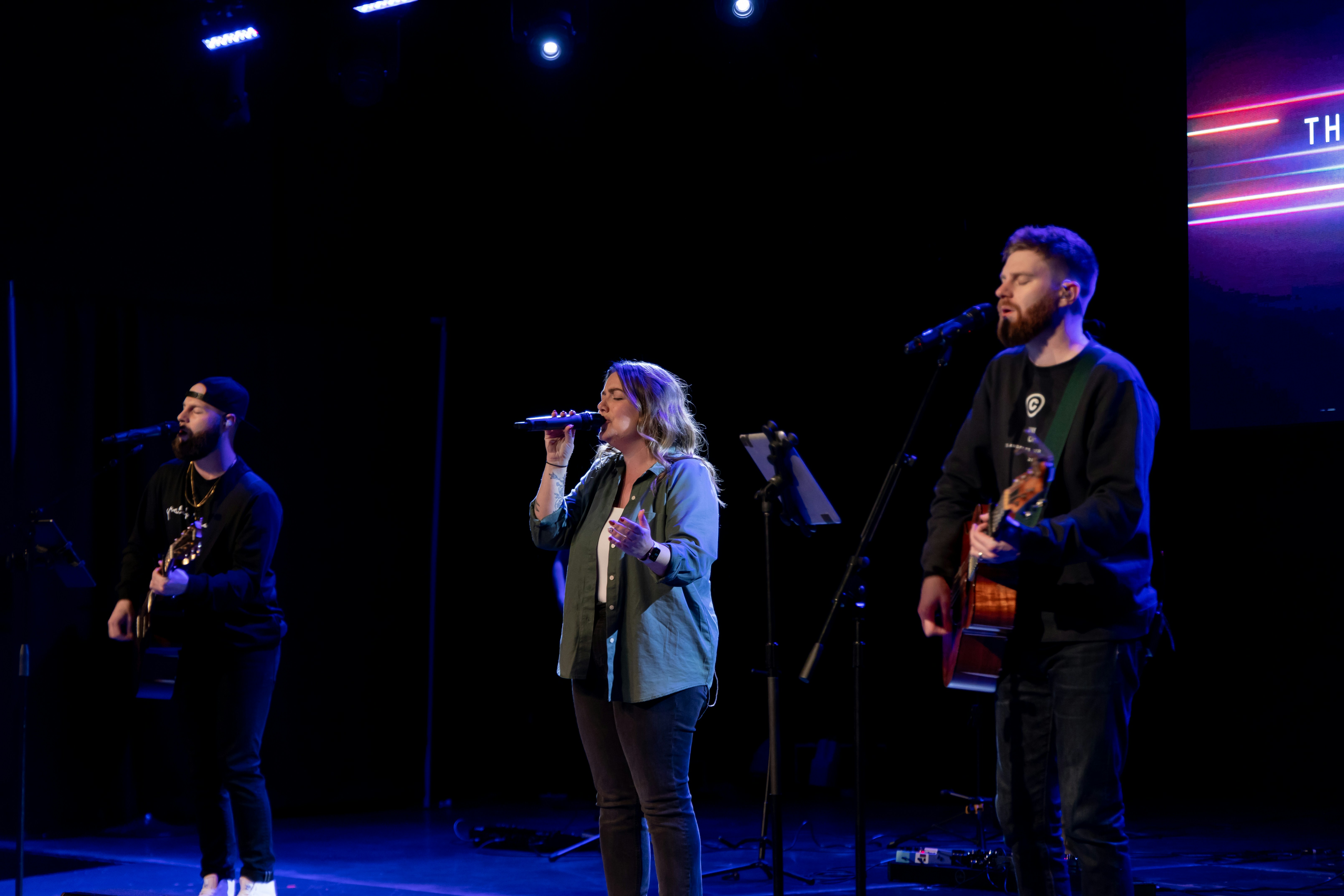 Three musicians perform on a dimly lit stage with guitars and microphones.