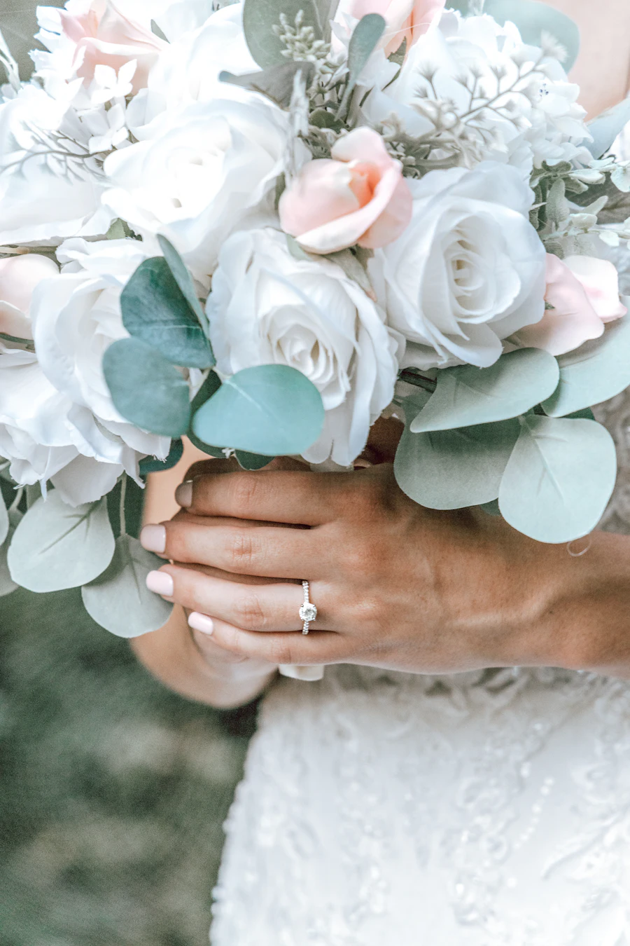 Close-up of a bride holding a bouquet of white roses and pale blush flowers with eucalyptus leaves, her hands showing a diamond ring. Soft, airy tones and a shallow depth of field create a romantic feel.