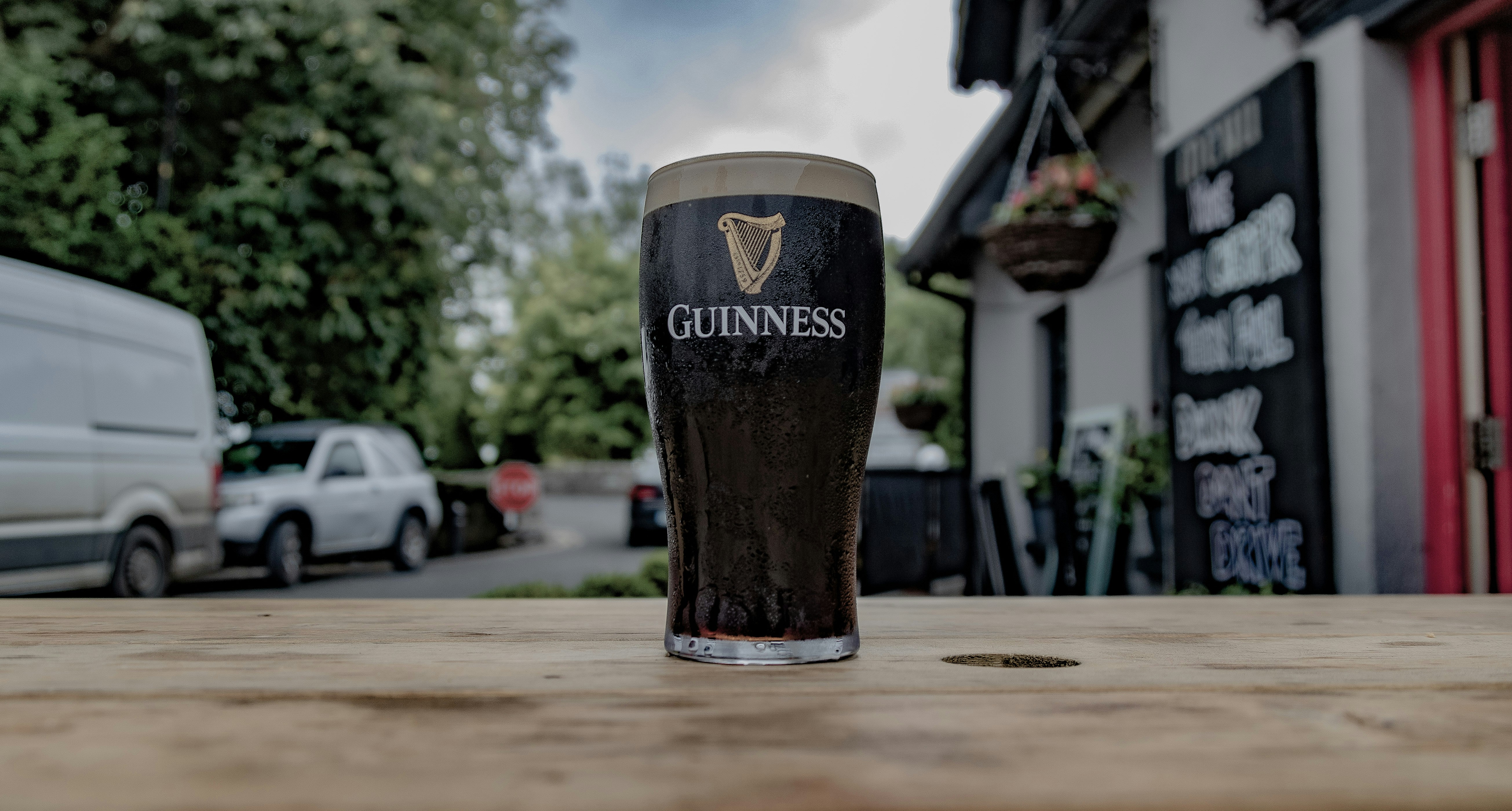 Pint of Guinness resting on a wooden table, with a lively pub atmosphere in the background. The iconic logo is prominently displayed.