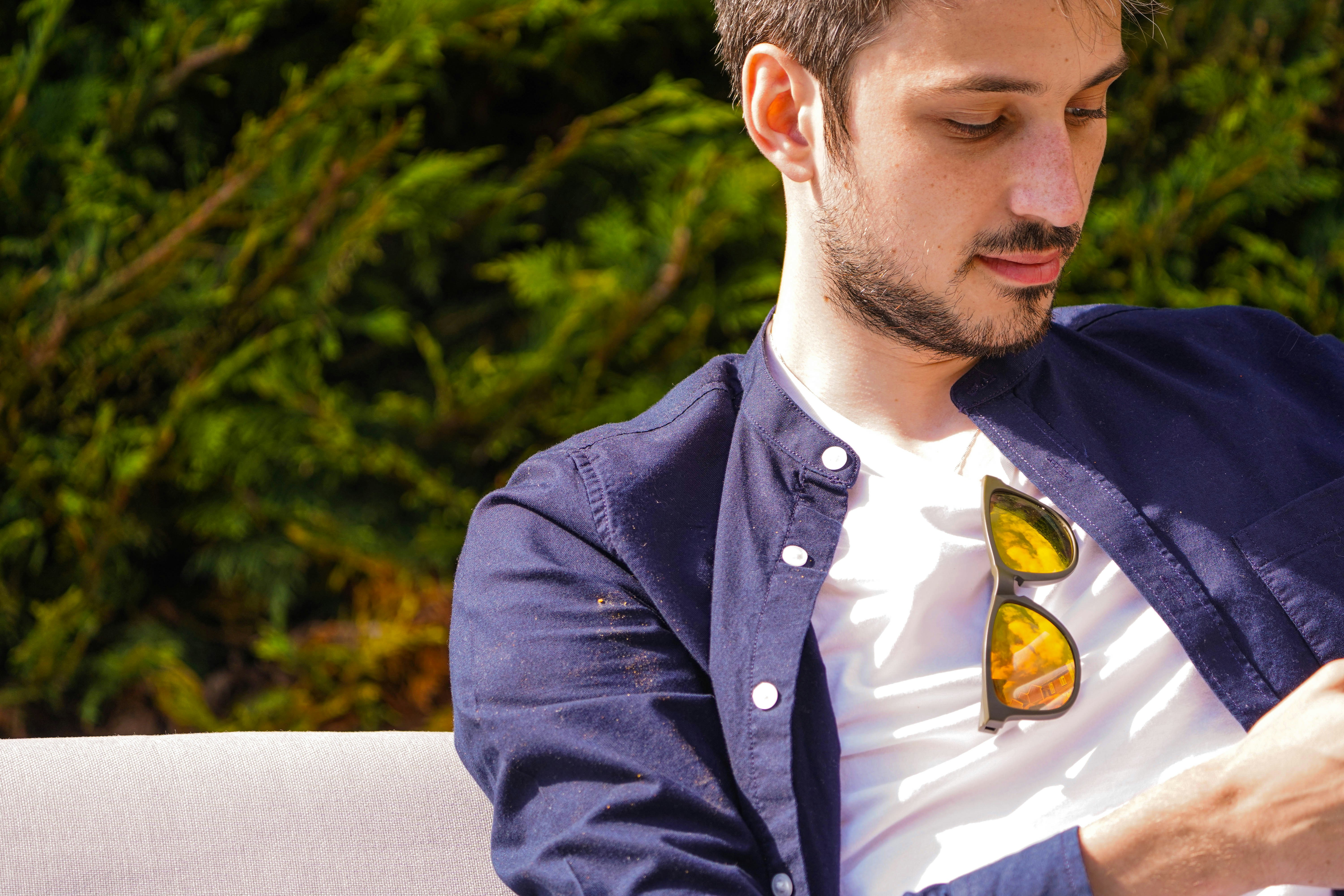 A young man seated outdoors, engrossed in his phone, with vibrant greenery framing the scene. Sunglasses rest on his shirt, adding a touch of casual style.