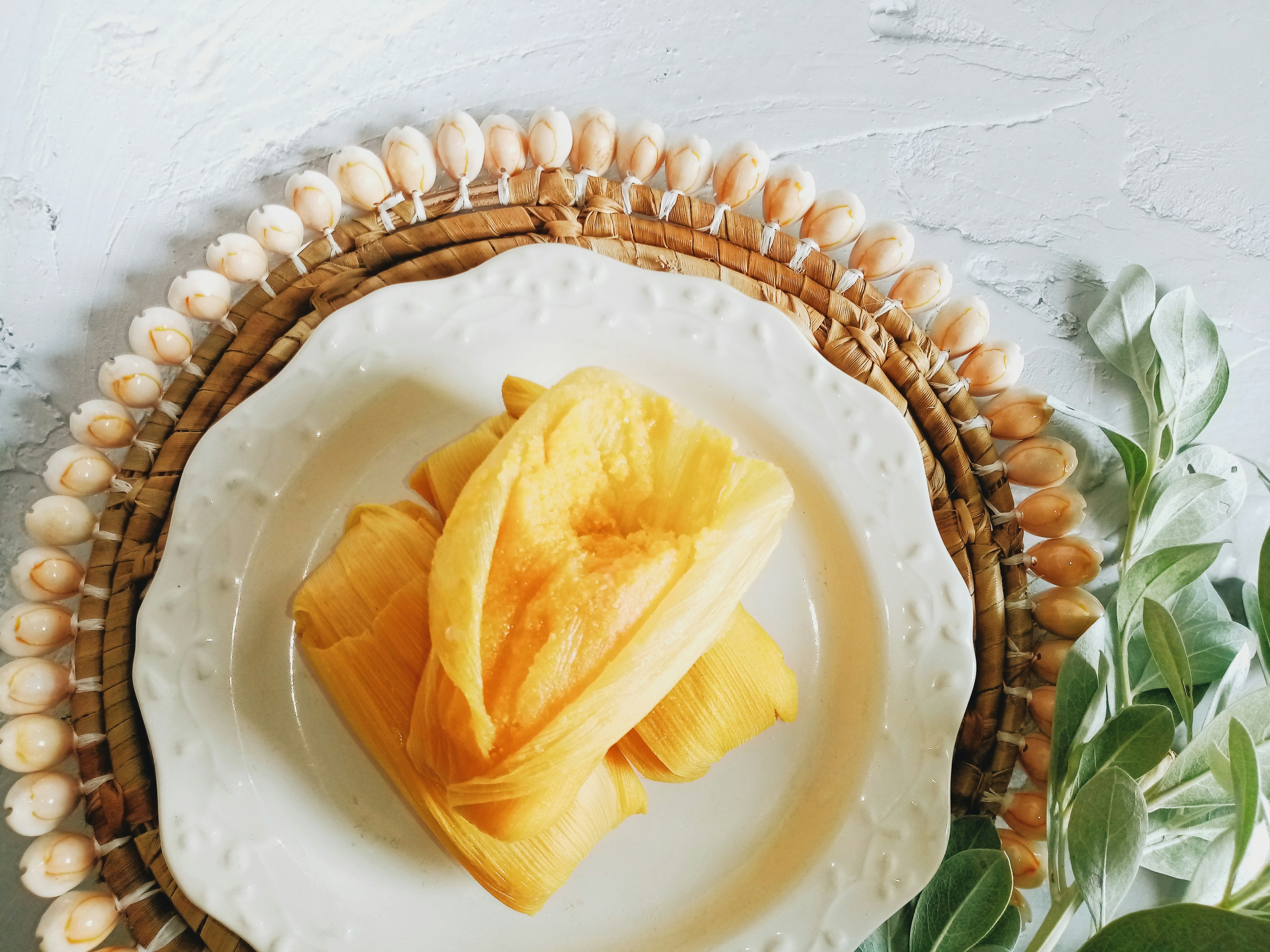 Yellow tamales neatly arranged on an elegant white plate, surrounded by a woven mat and green foliage.