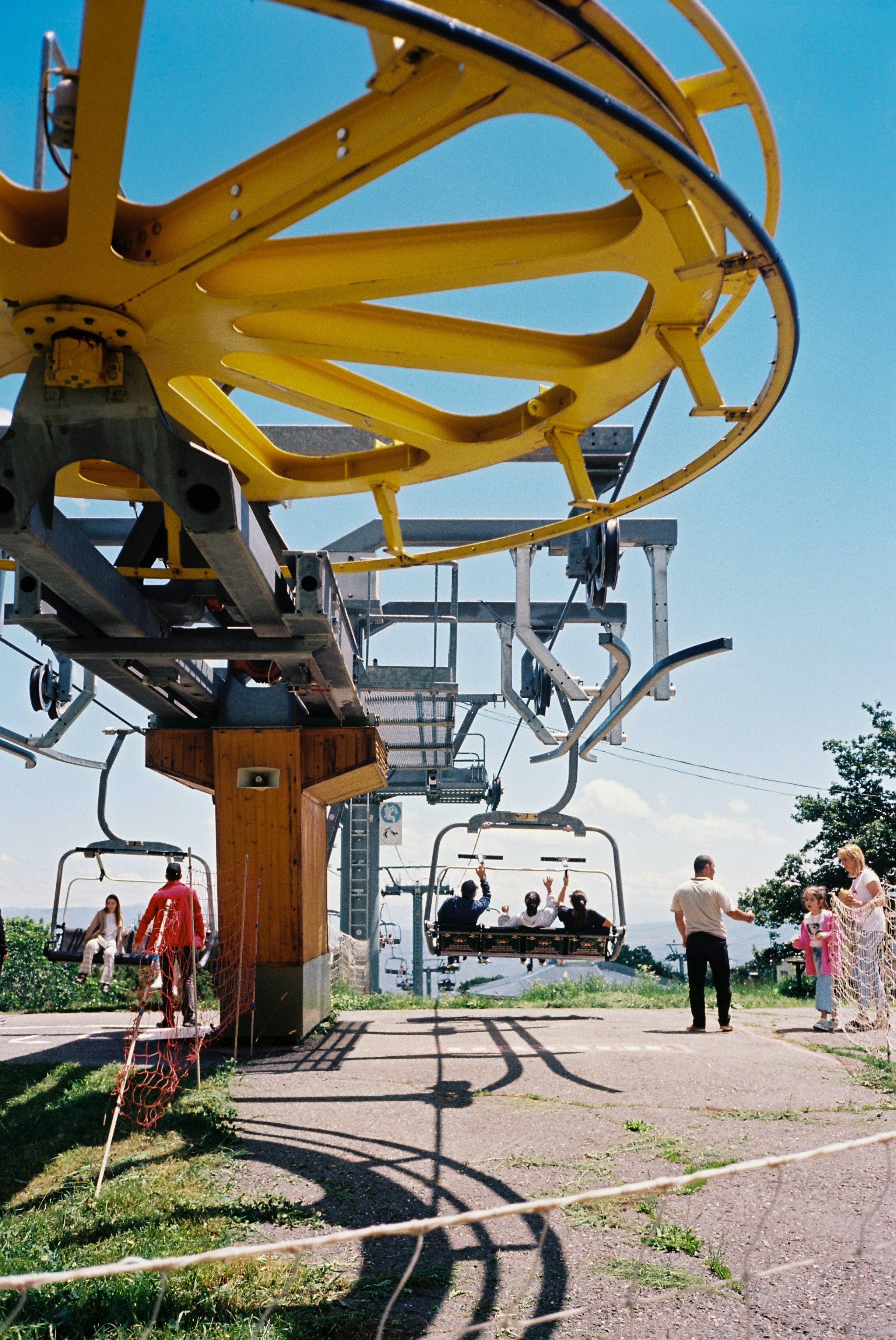 A chairlift station bustling with visitors, featuring a bright yellow lift mechanism against a clear blue sky. The scene captures the anticipation of adventure and leisure.
