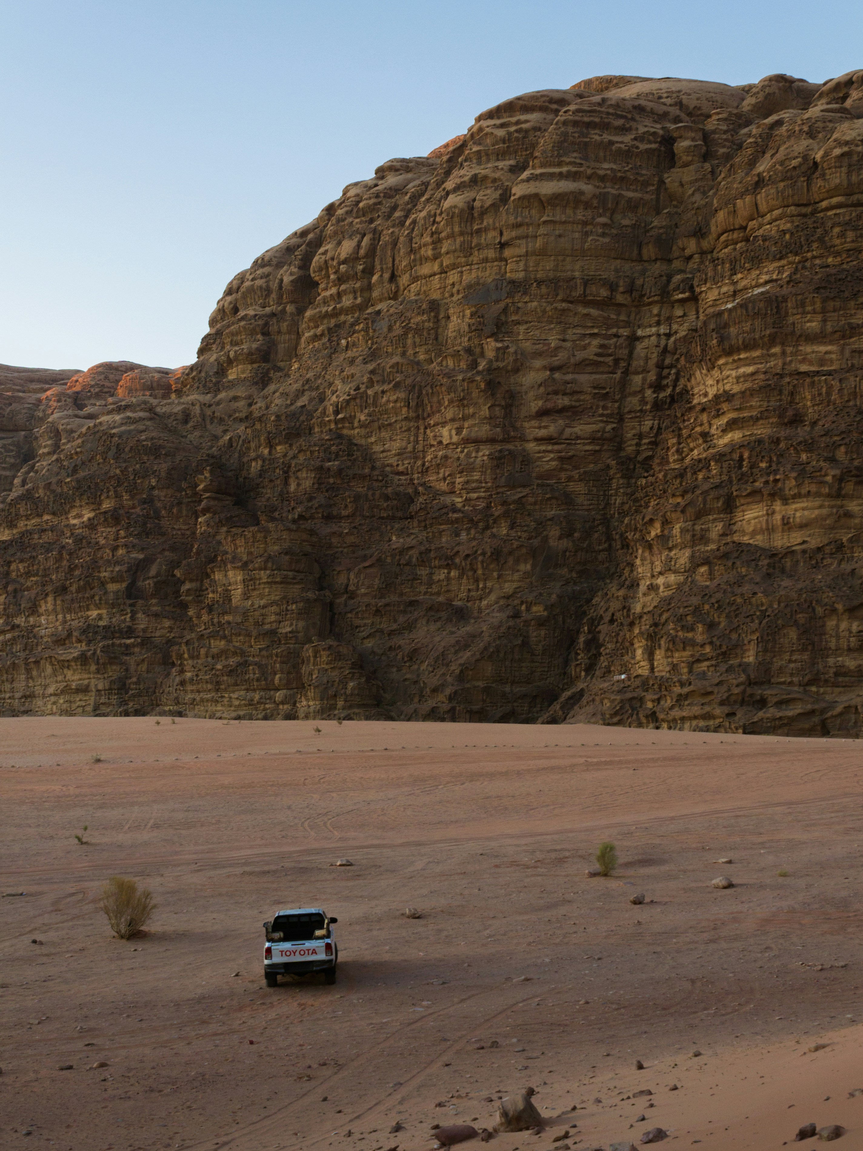 A lone vehicle parked in a vast desert landscape with towering sandstone cliffs under a clear sky.