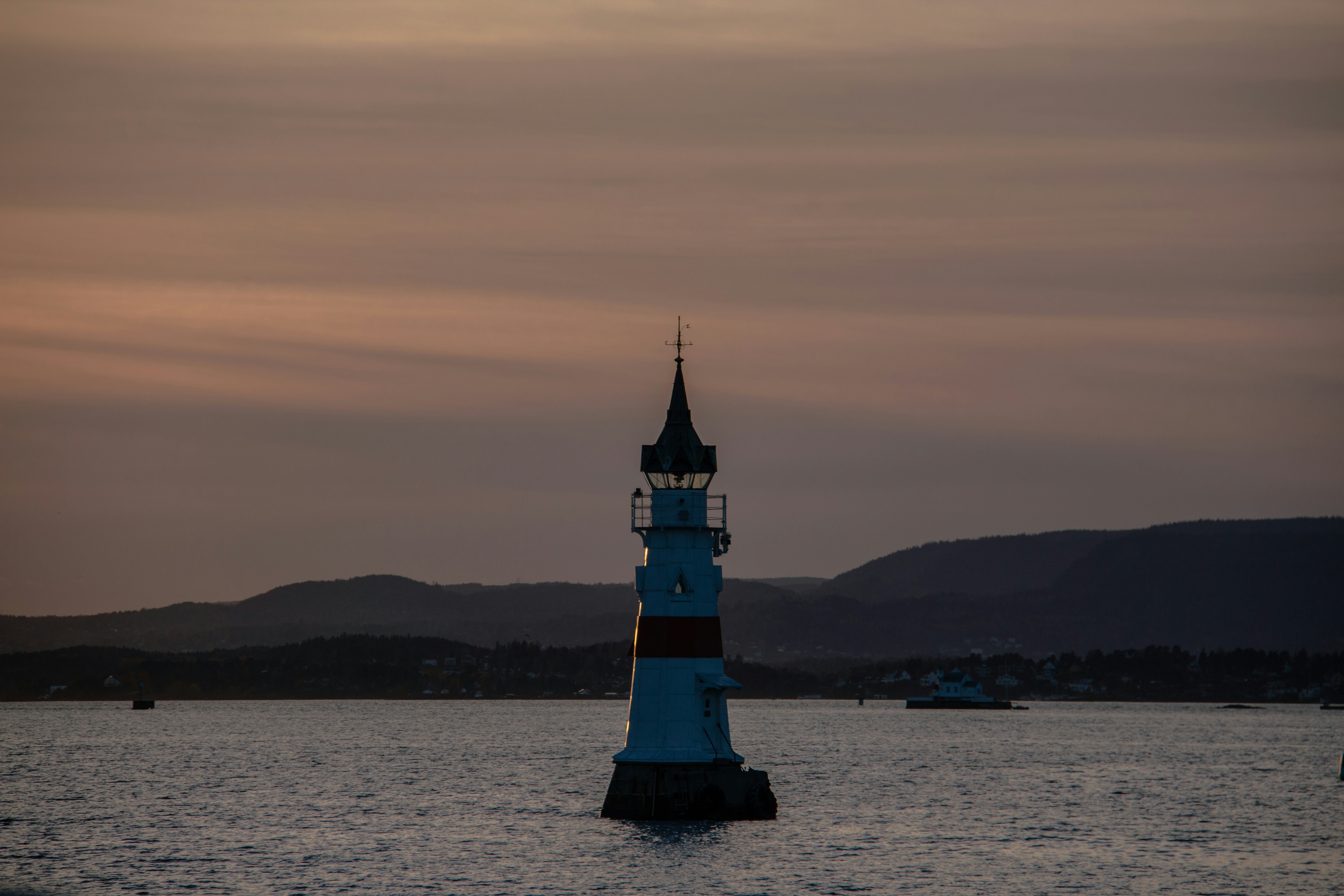 Lighthouse standing tall in calm waters against a softly lit twilight sky, silhouetted by distant hills.