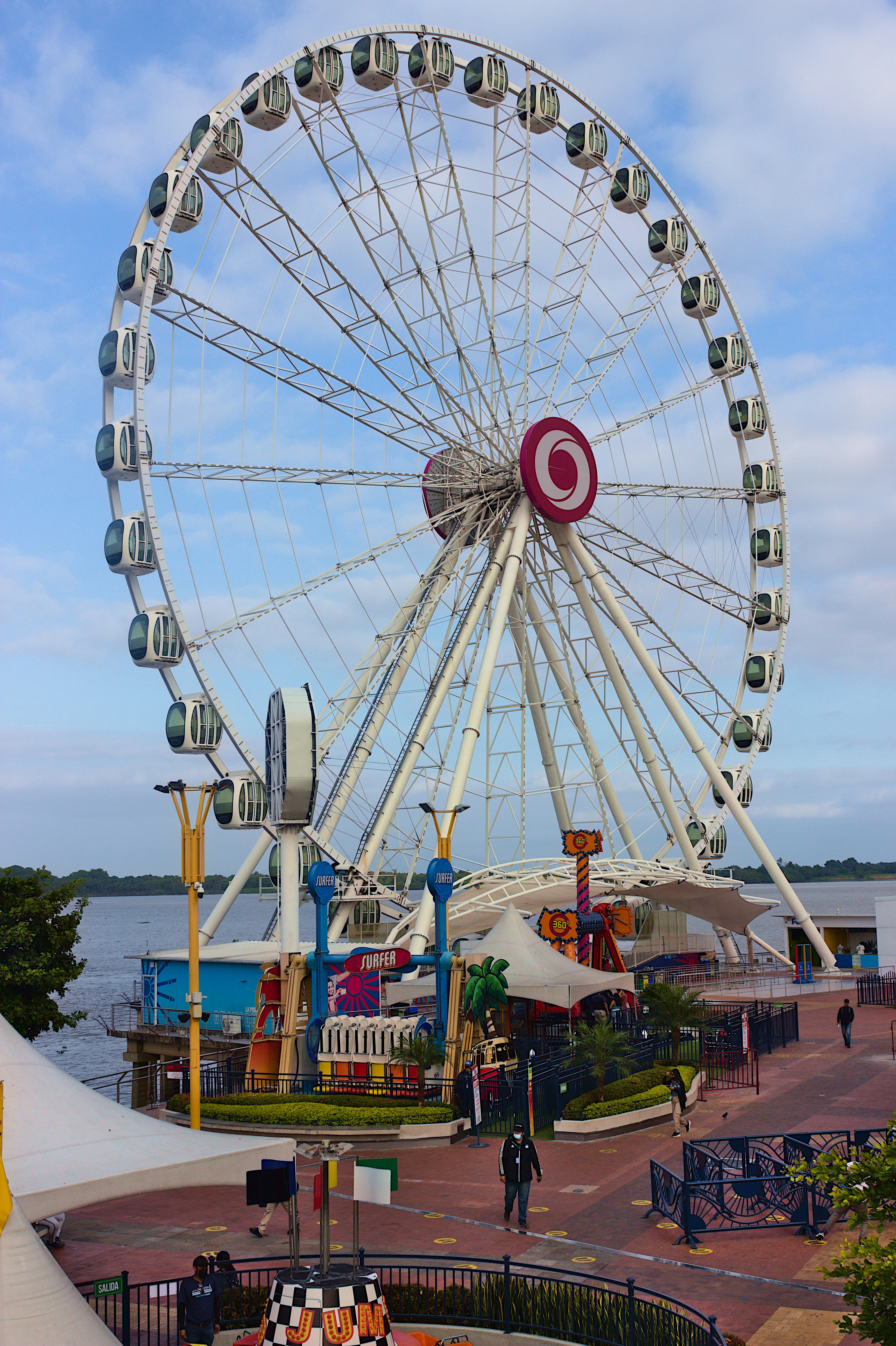 A vibrant Ferris wheel stands tall against a cloudy sky, surrounded by amusement park attractions and a serene waterfront. The scene captures the essence of fun and leisure.