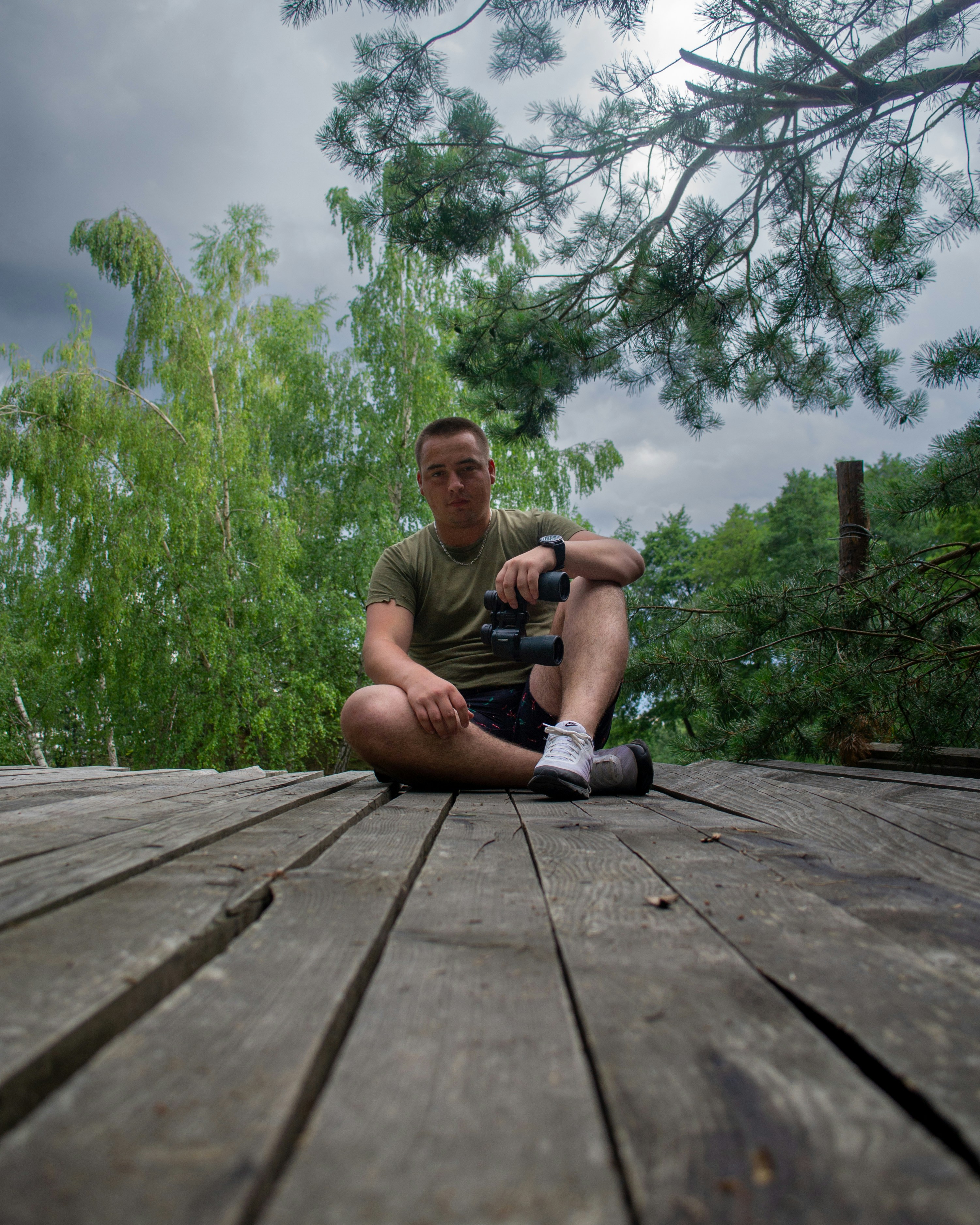 Individual seated on a wooden deck surrounded by lush greenery and overcast skies, holding a camera. The scene conveys a blend of solitude and connection with nature.