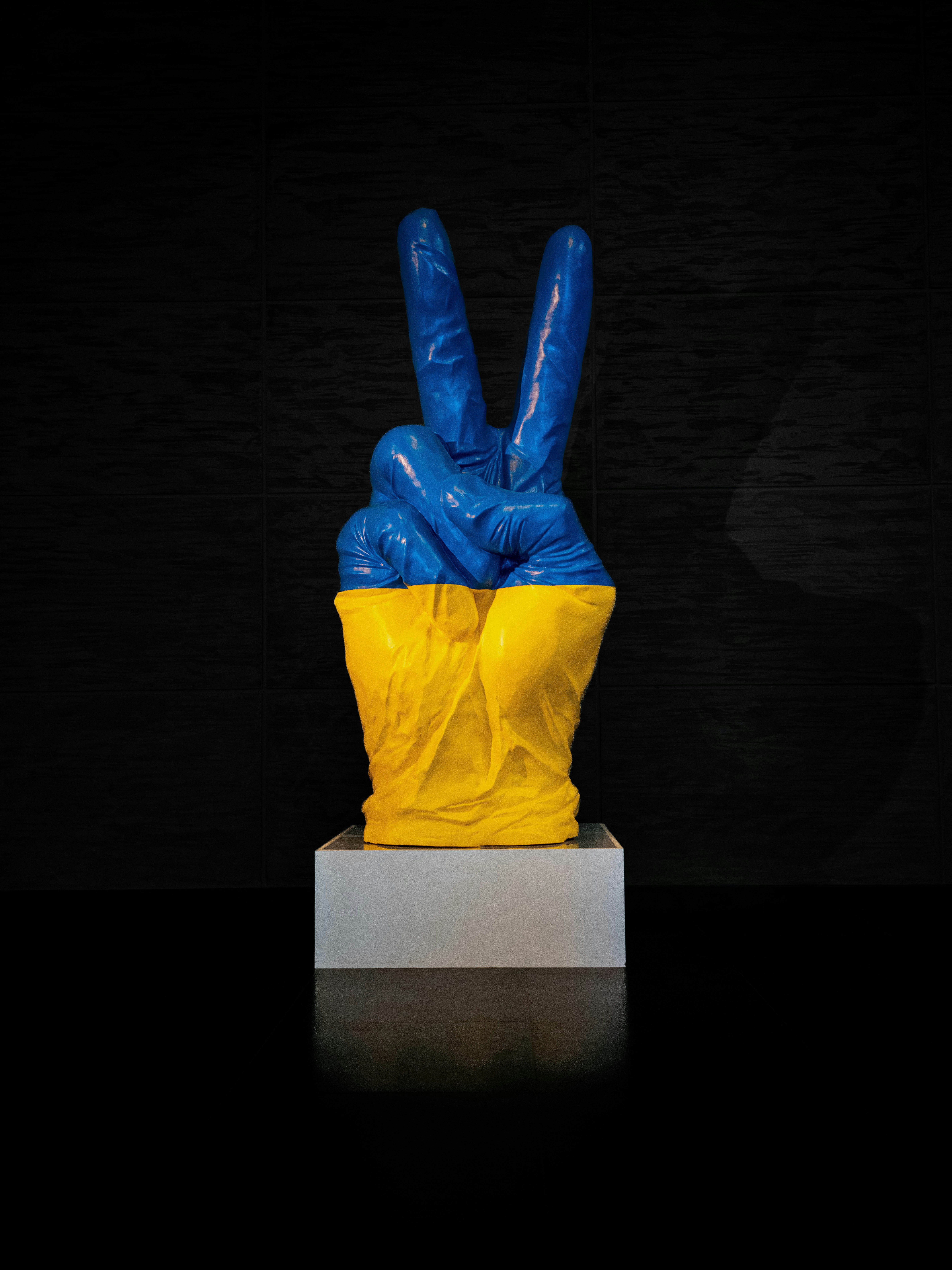 A large sculpture of a peace sign hand, colored in blue and yellow, symbolizing hope and unity. The artwork is displayed prominently against a dark background.