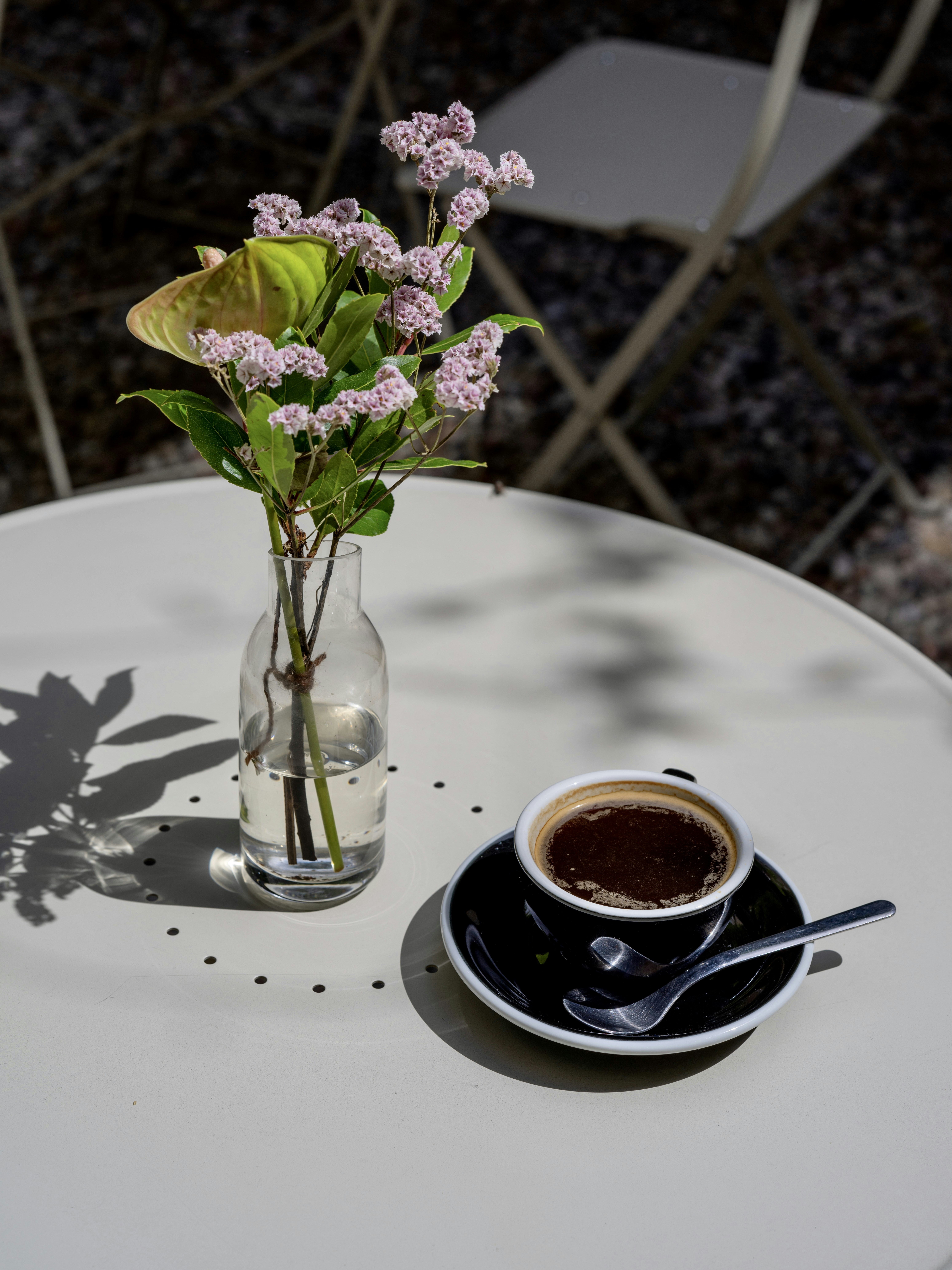 A delicate vase of flowers beside a steaming cup of coffee on a round table, casting soft shadows in natural light.
