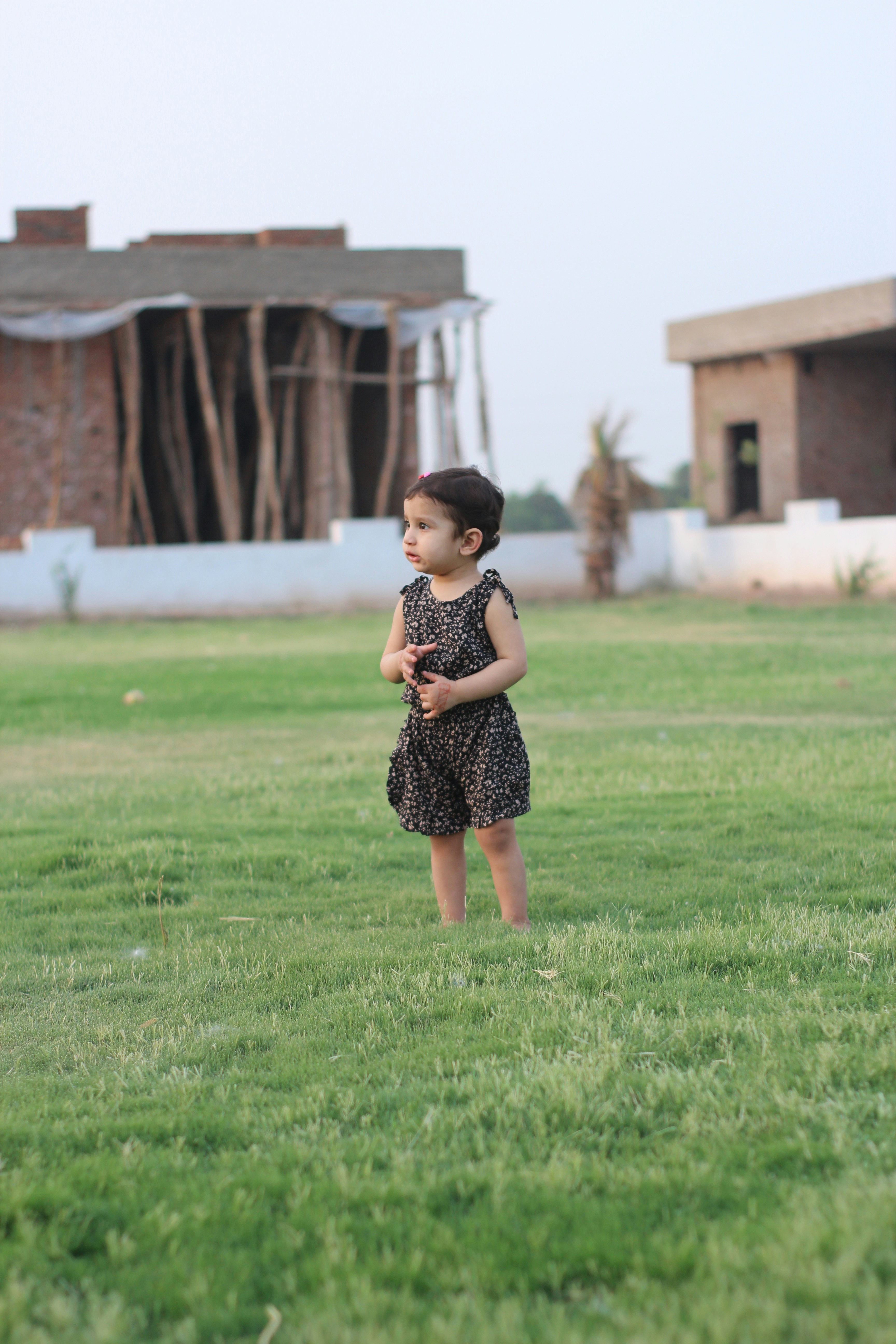 A young child stands in a lush green field, gazing thoughtfully while holding a small object. Abandoned structures loom in the background, adding depth to the scene.