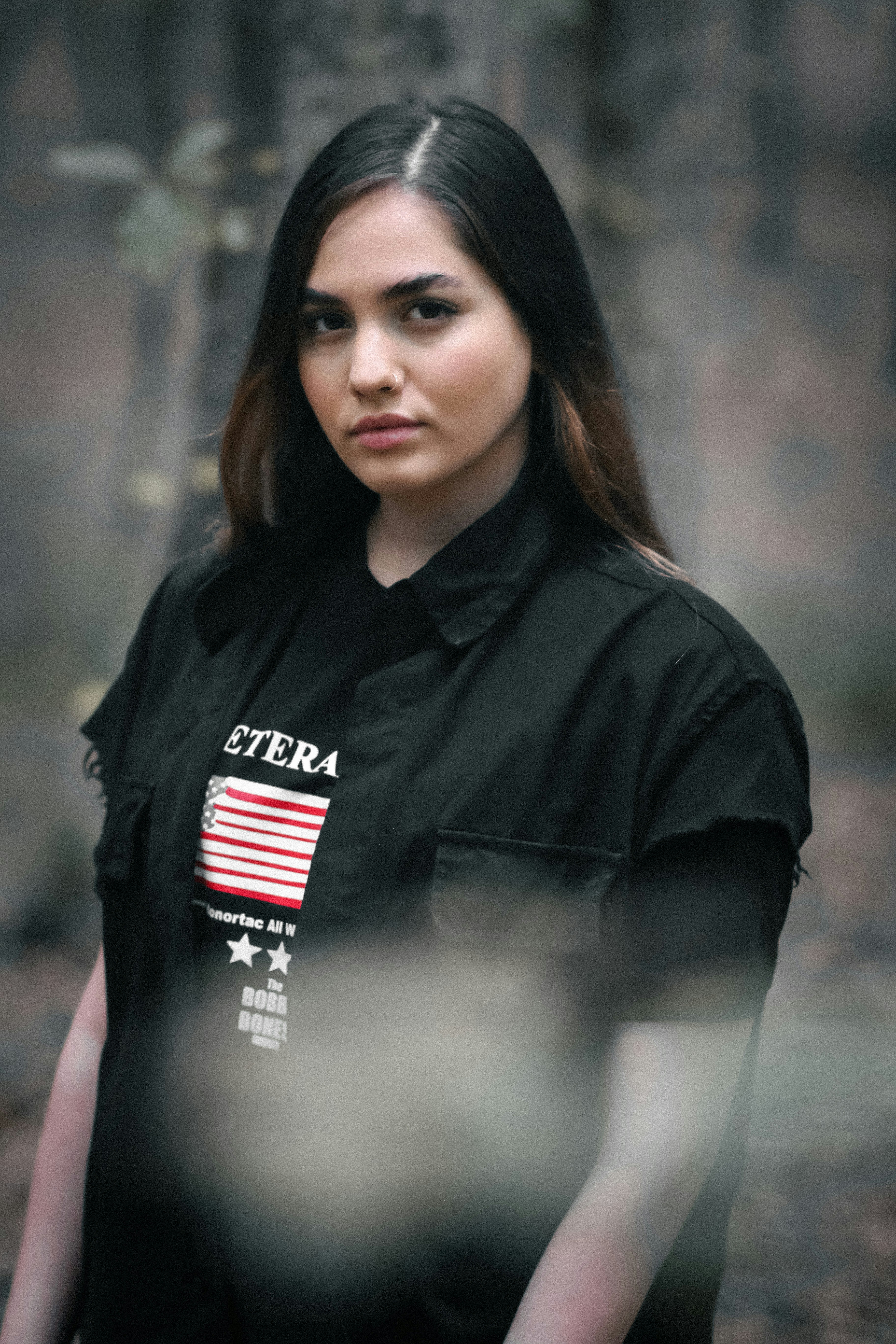 A young woman stands confidently in a forest, wearing a black shirt with a patriotic design. The background is softly blurred, emphasizing her presence.