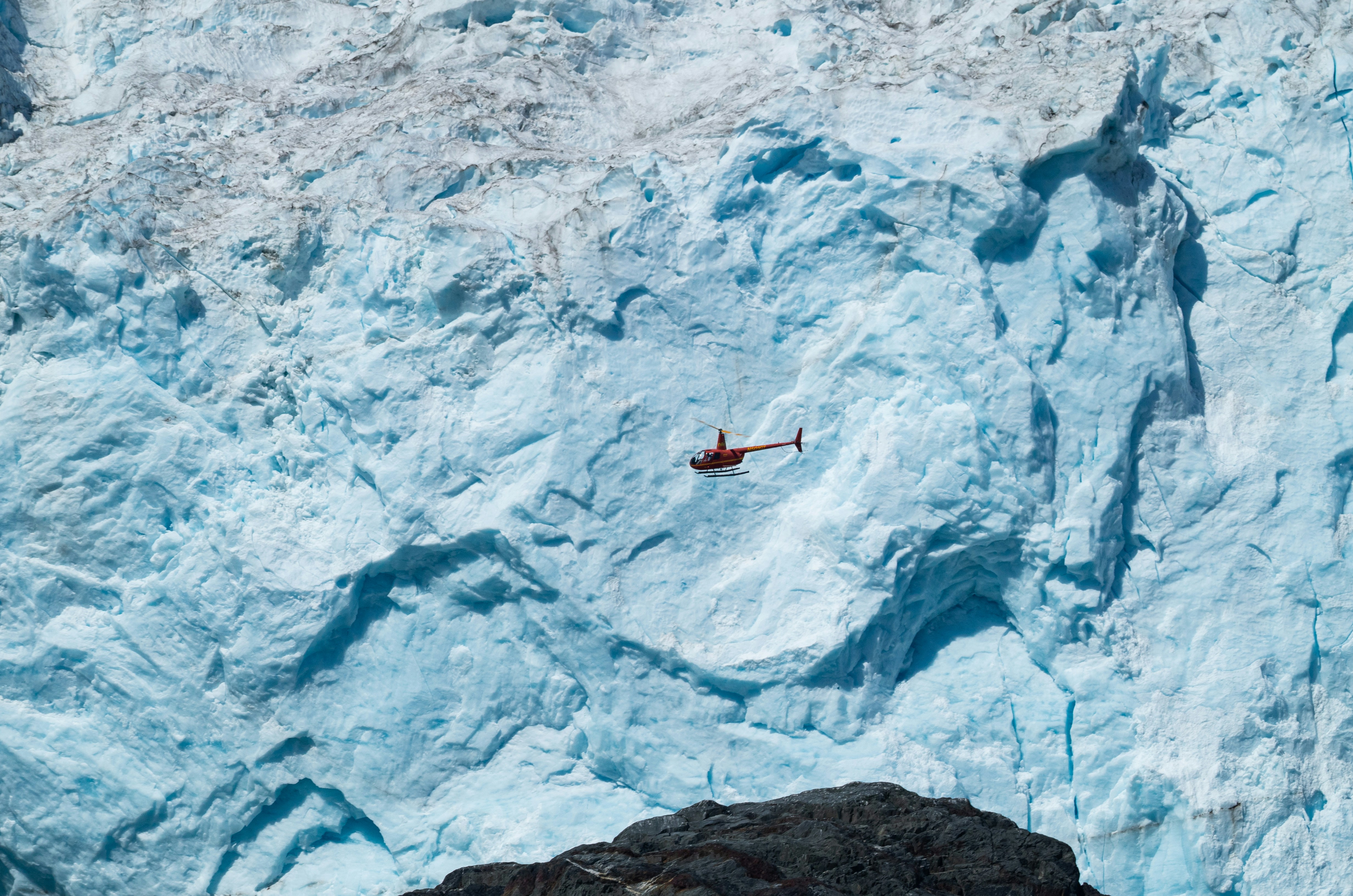 A red helicopter hovers near a vast, textured glacier wall, emphasizing the scale of the ice formation.