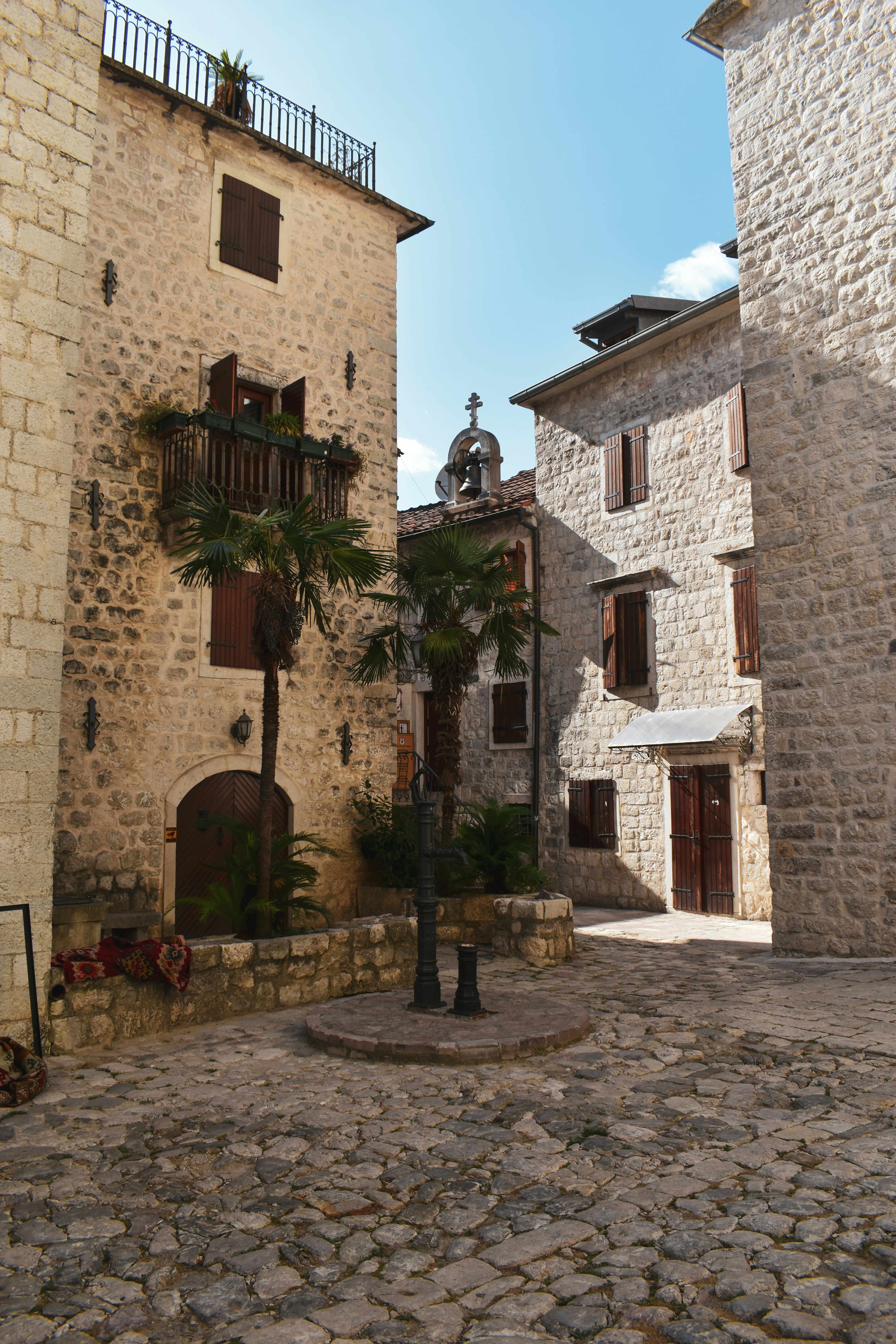 Charming cobbled square surrounded by rustic stone buildings with wooden shutters and palm trees. A vintage lamp post adds character to the serene atmosphere.