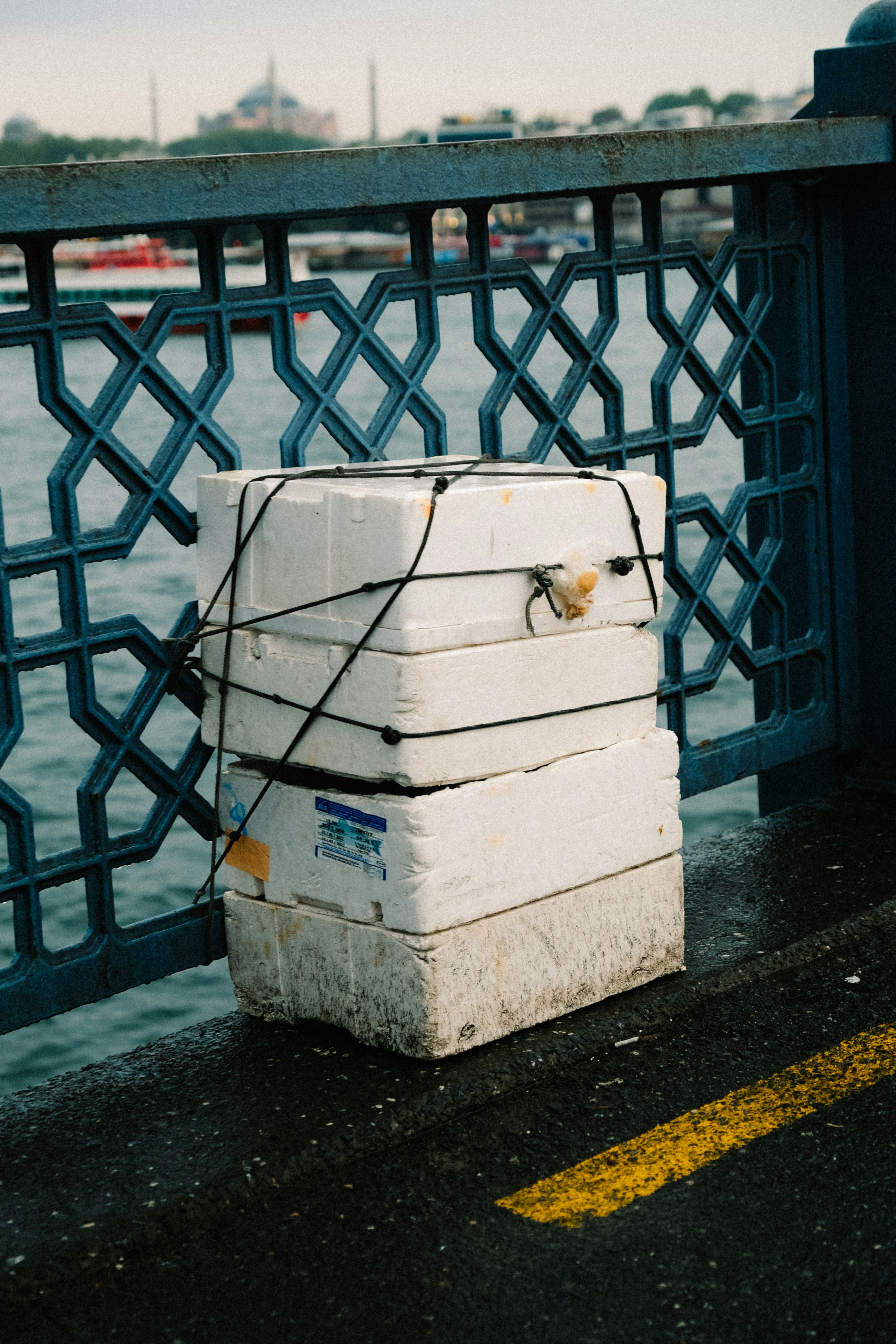 Three stacked white coolers secured with black straps against a blue railing, overlooking a calm body of water in an urban setting.