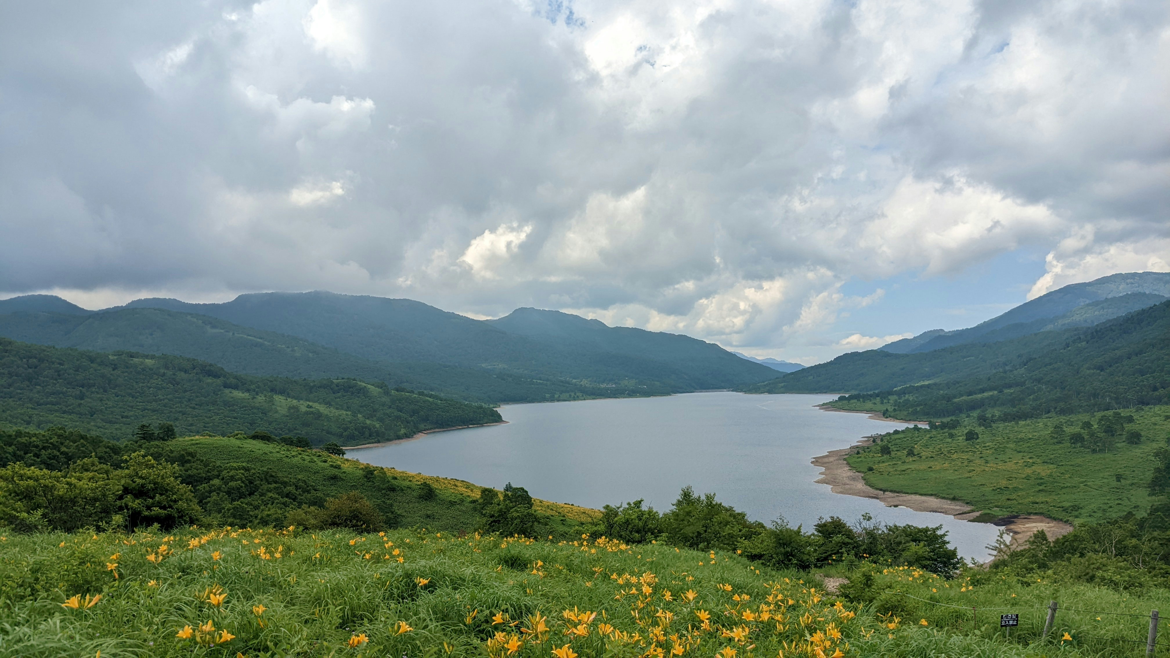 A panoramic photograph of a tranquil valley with a winding lake, green meadows dotted with yellow wildflowers, and distant cloud-covered mountains.