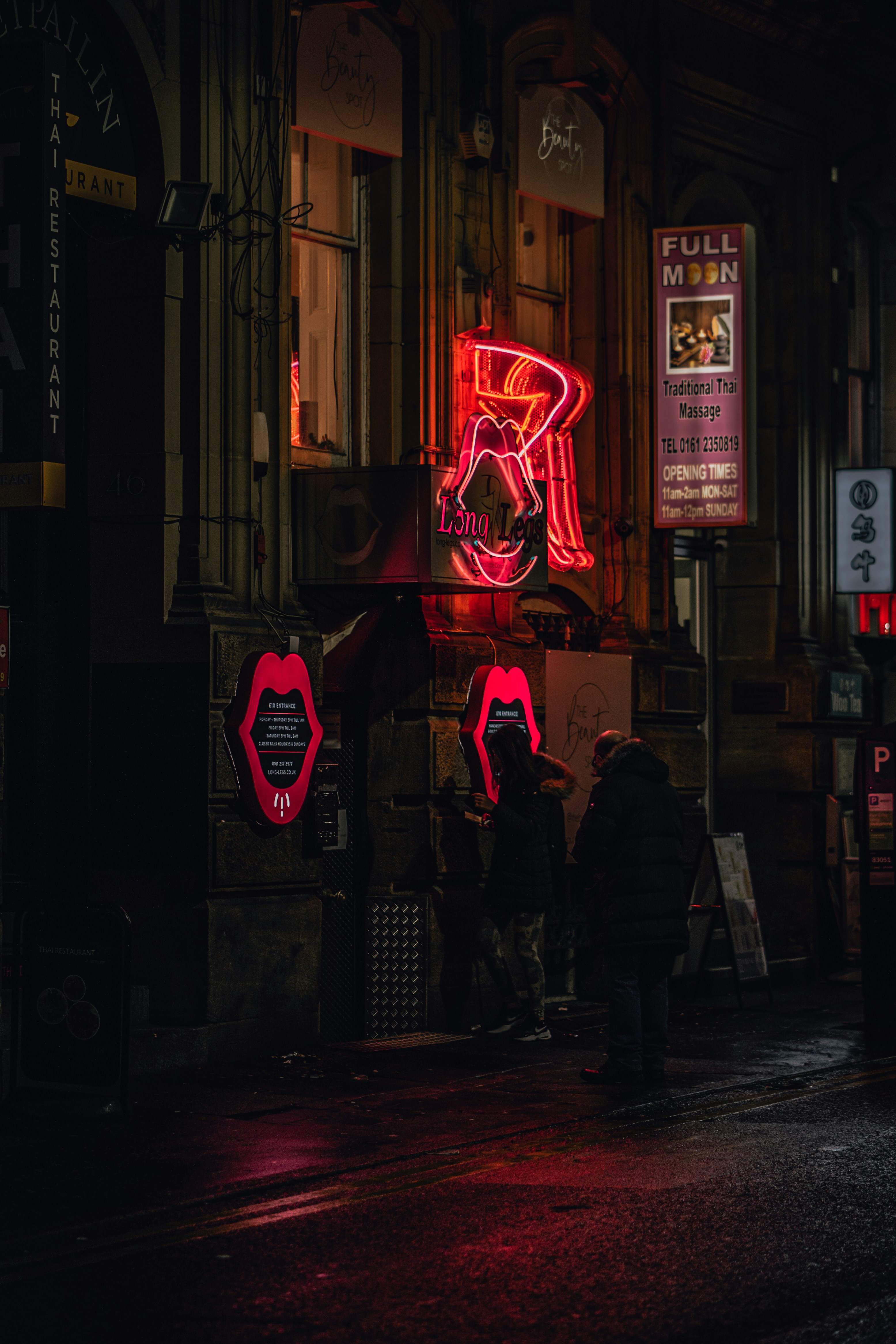 Vibrant neon signage illuminates a bustling street corner, highlighting two figures engaged in conversation under the glow of pink lips. The wet pavement reflects the colorful lights.