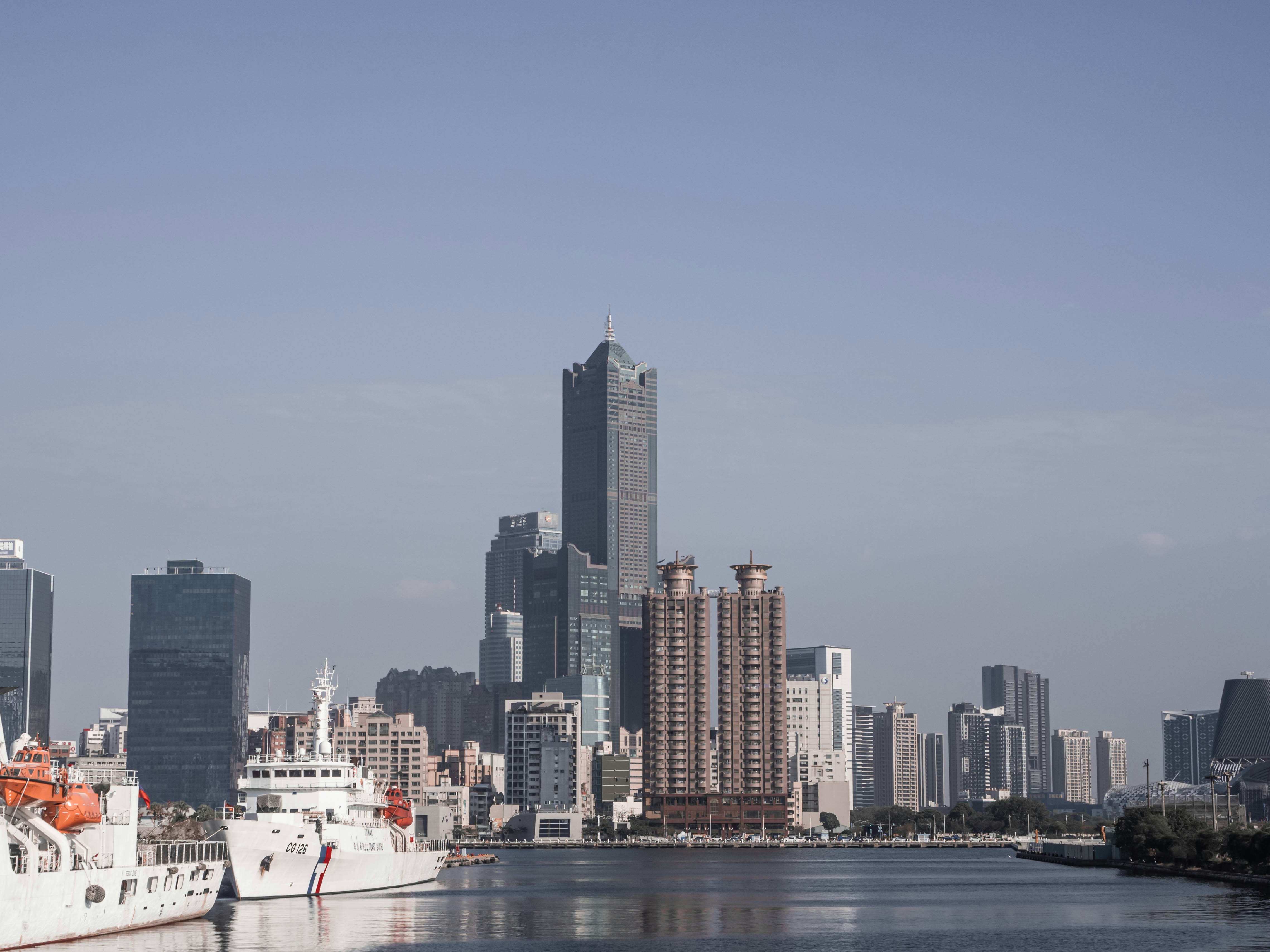 City skyline with towering buildings and a boat on calm waters under a clear blue sky.