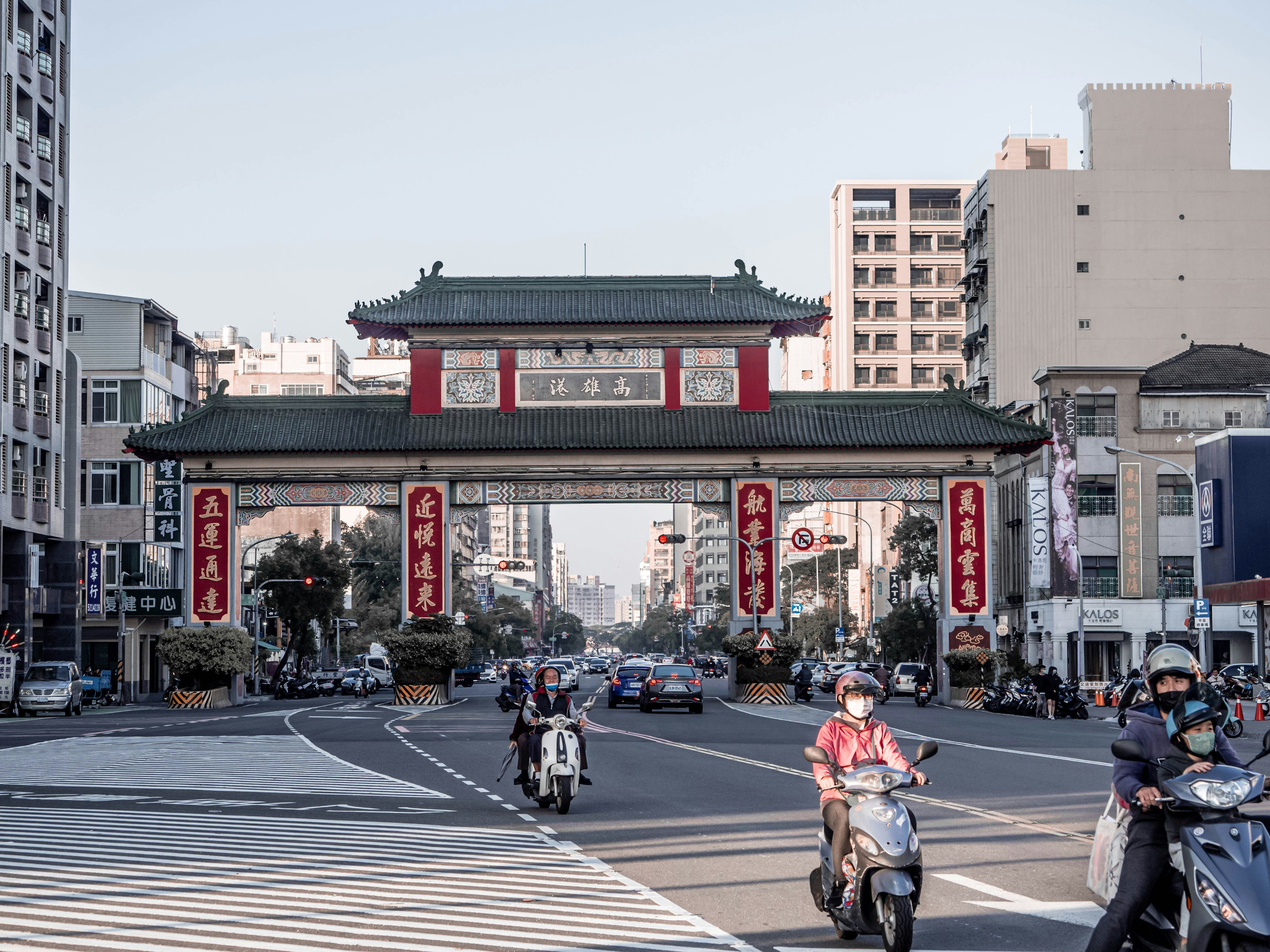 Motorcyclists traverse a busy city street beneath a traditional Chinese archway surrounded by modern buildings.