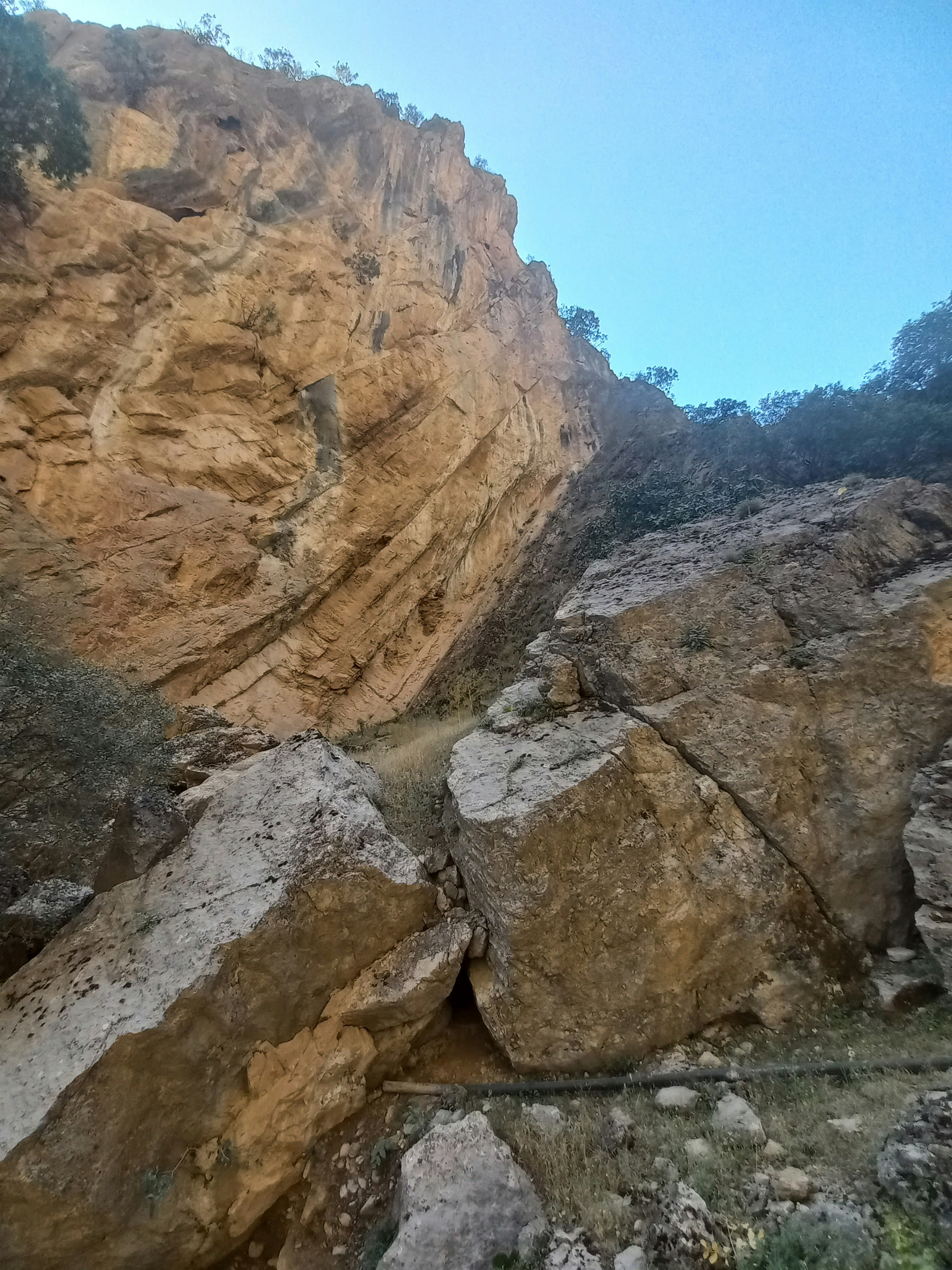 Imposing rocky cliffside towers over a rugged terrain, with scattered boulders and sparse vegetation below. The clear blue sky contrasts sharply with the earthy tones of the rock formations.