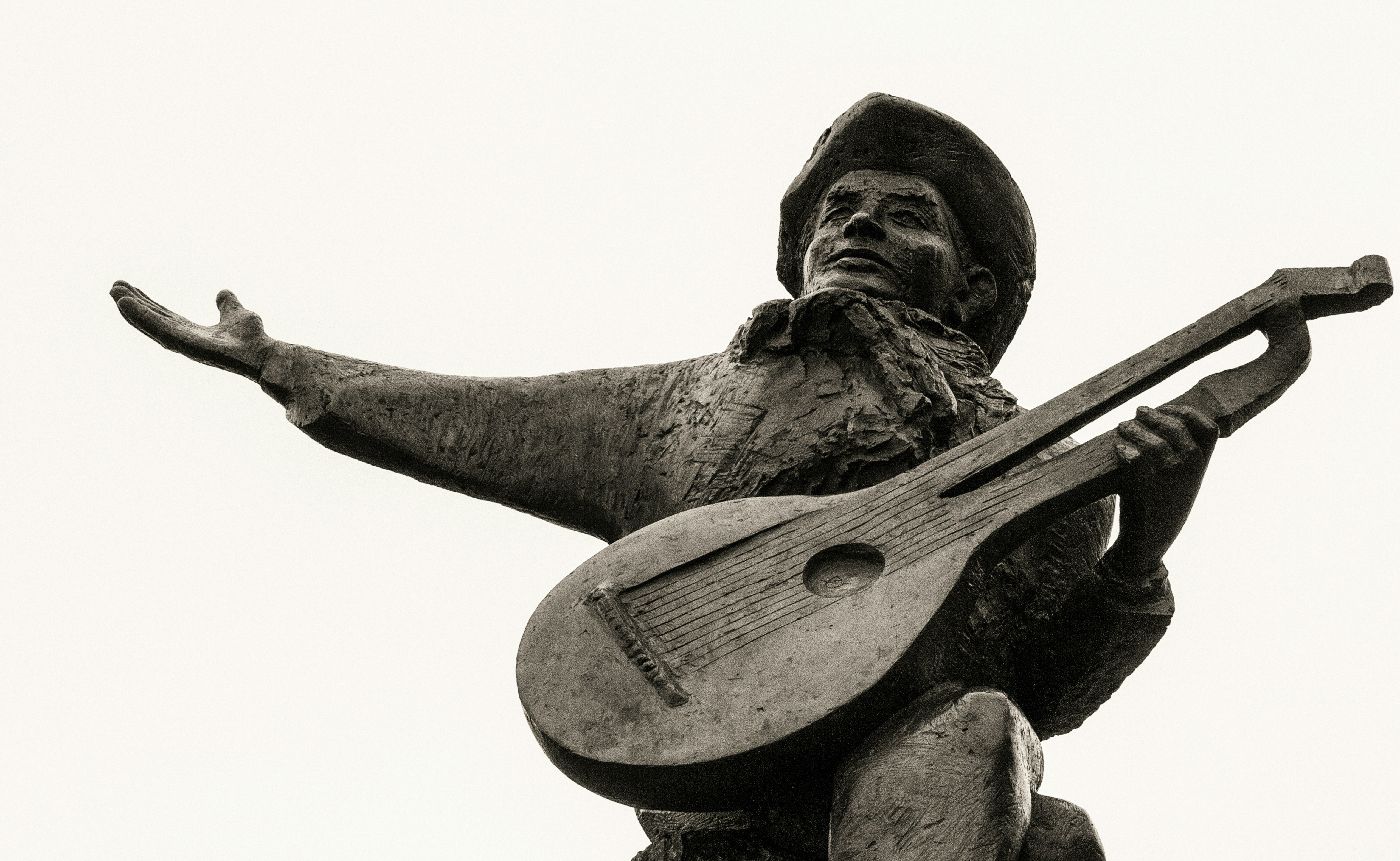 A weathered bronze statue of a musician with arms outstretched against a pale sky. The sculpture highlights a rounded, lute-like instrument and the performer's wide, expressive pose.