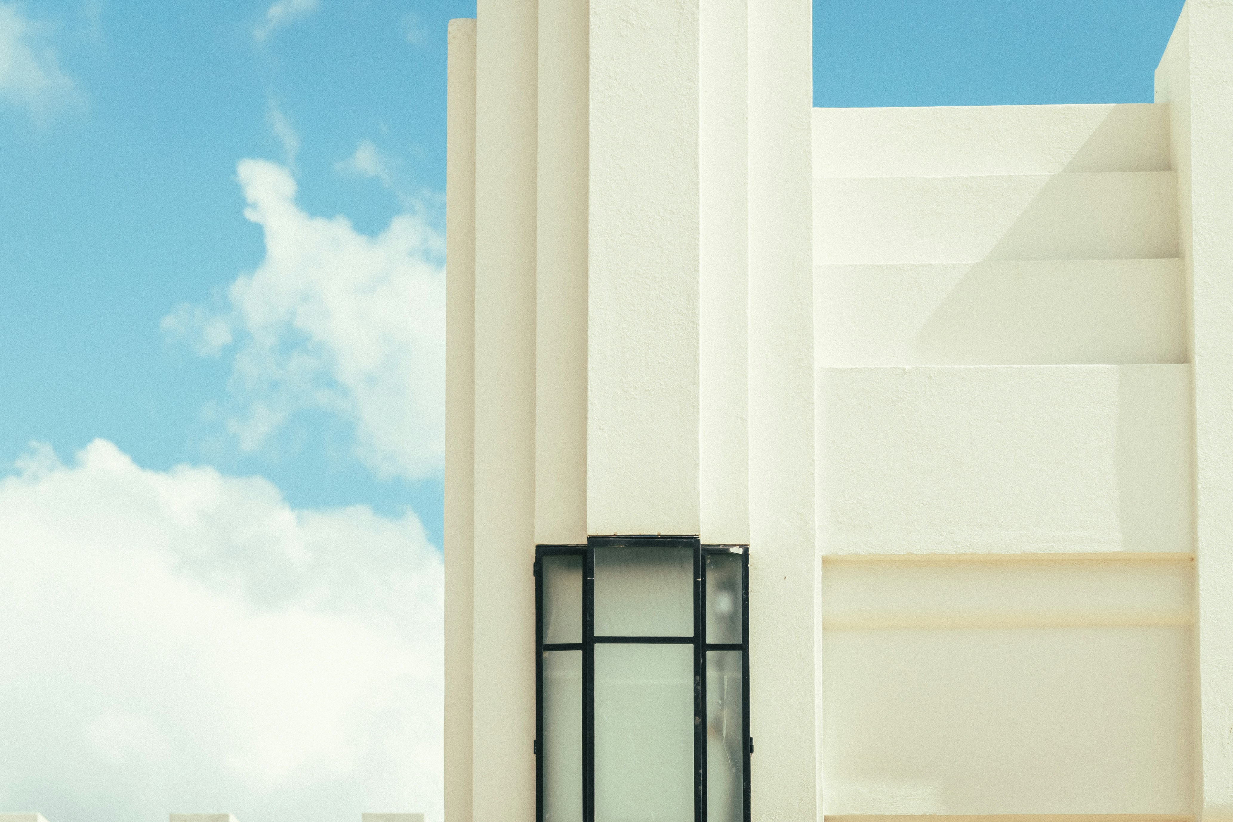 Modern architectural detail featuring a white facade and large glass window against a bright blue sky.